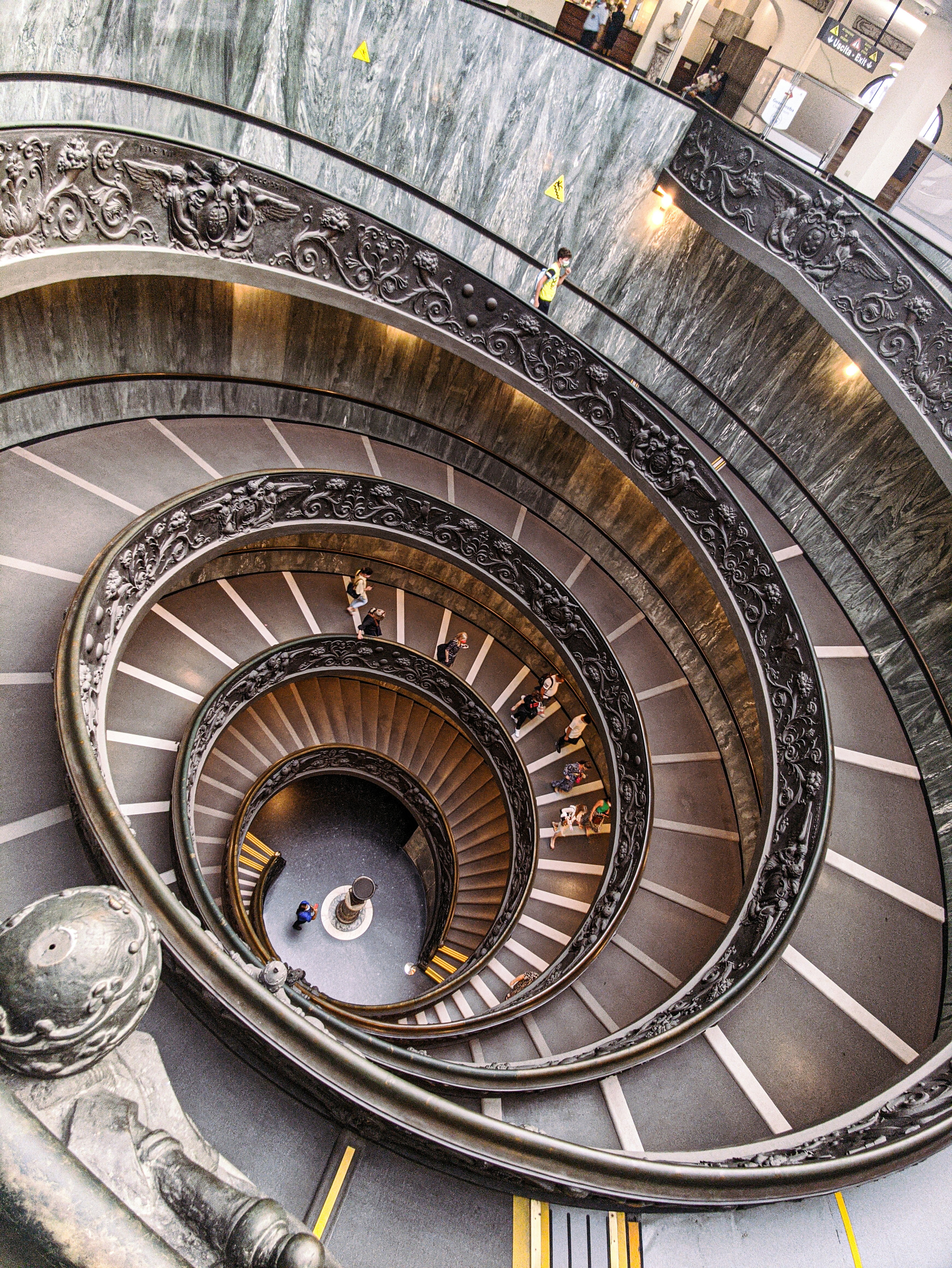 Intricate spiral staircase with ornate railings, leading down to a central point where visitors gather.