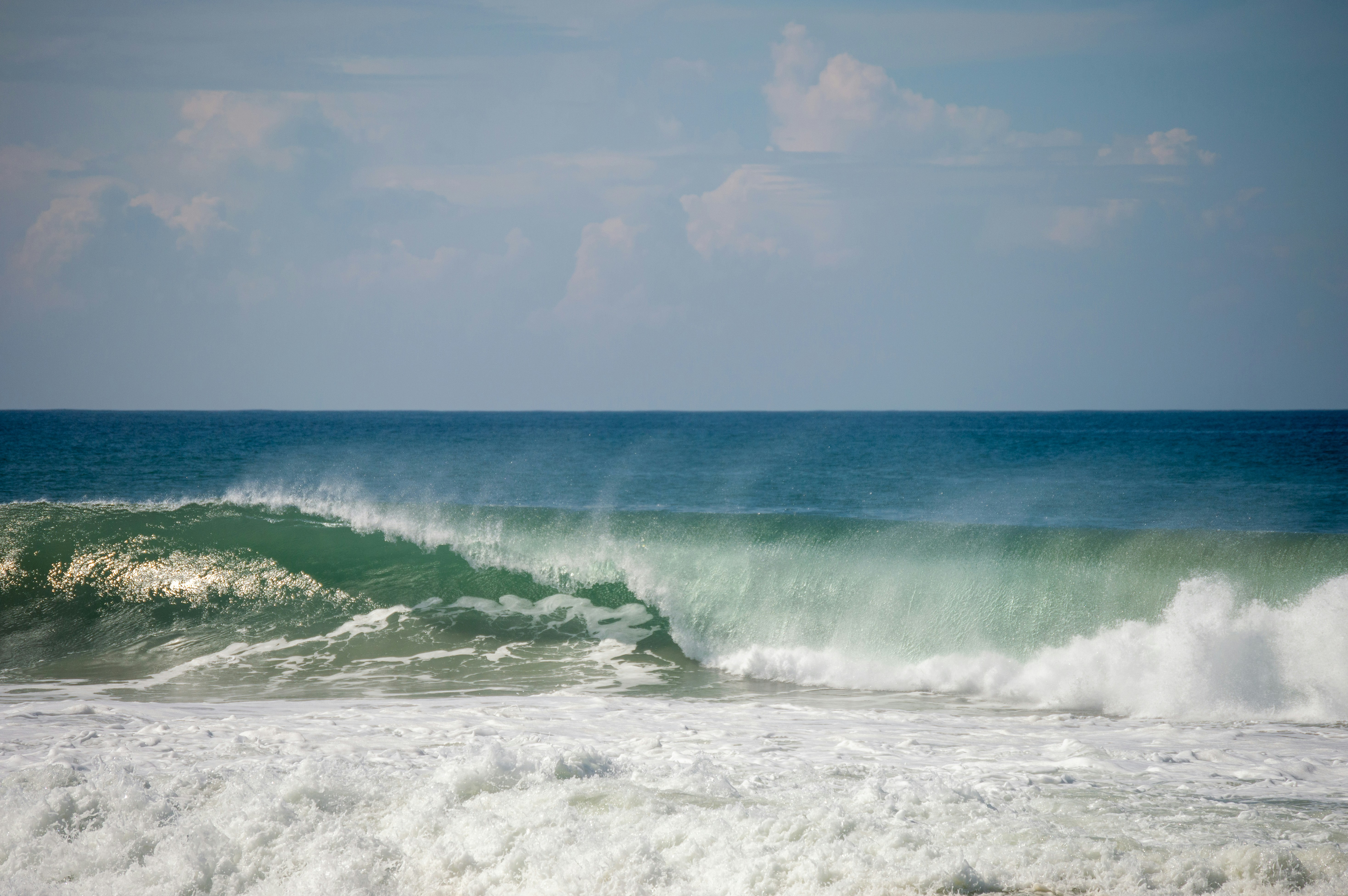Dynamic wave curling at the shoreline, with sunlight glinting off its surface and a backdrop of serene blue skies.