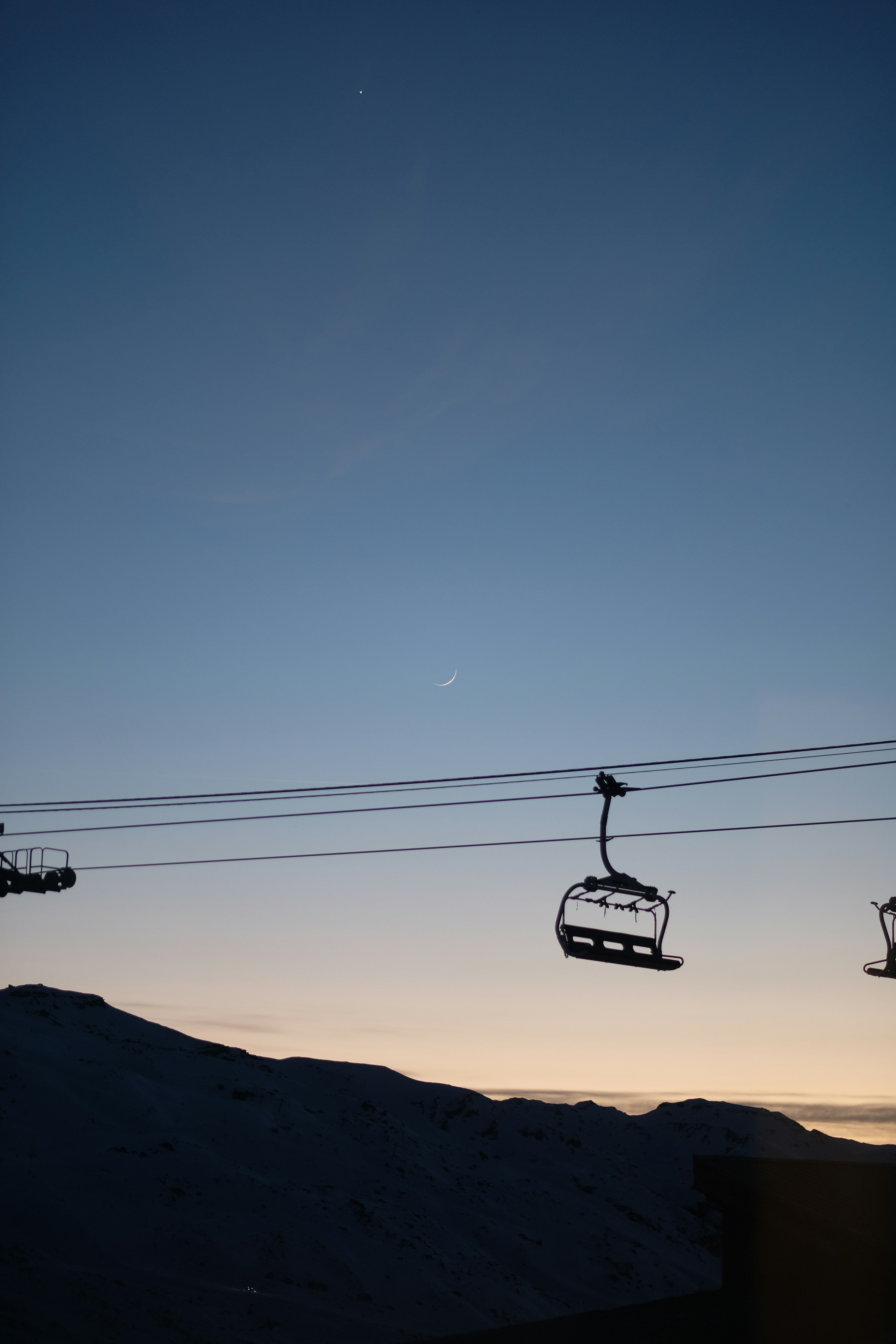 Ski lift chairs silhouetted against a twilight sky, with a crescent moon and a distant star visible. The serene ambiance captures the transition from day to night.