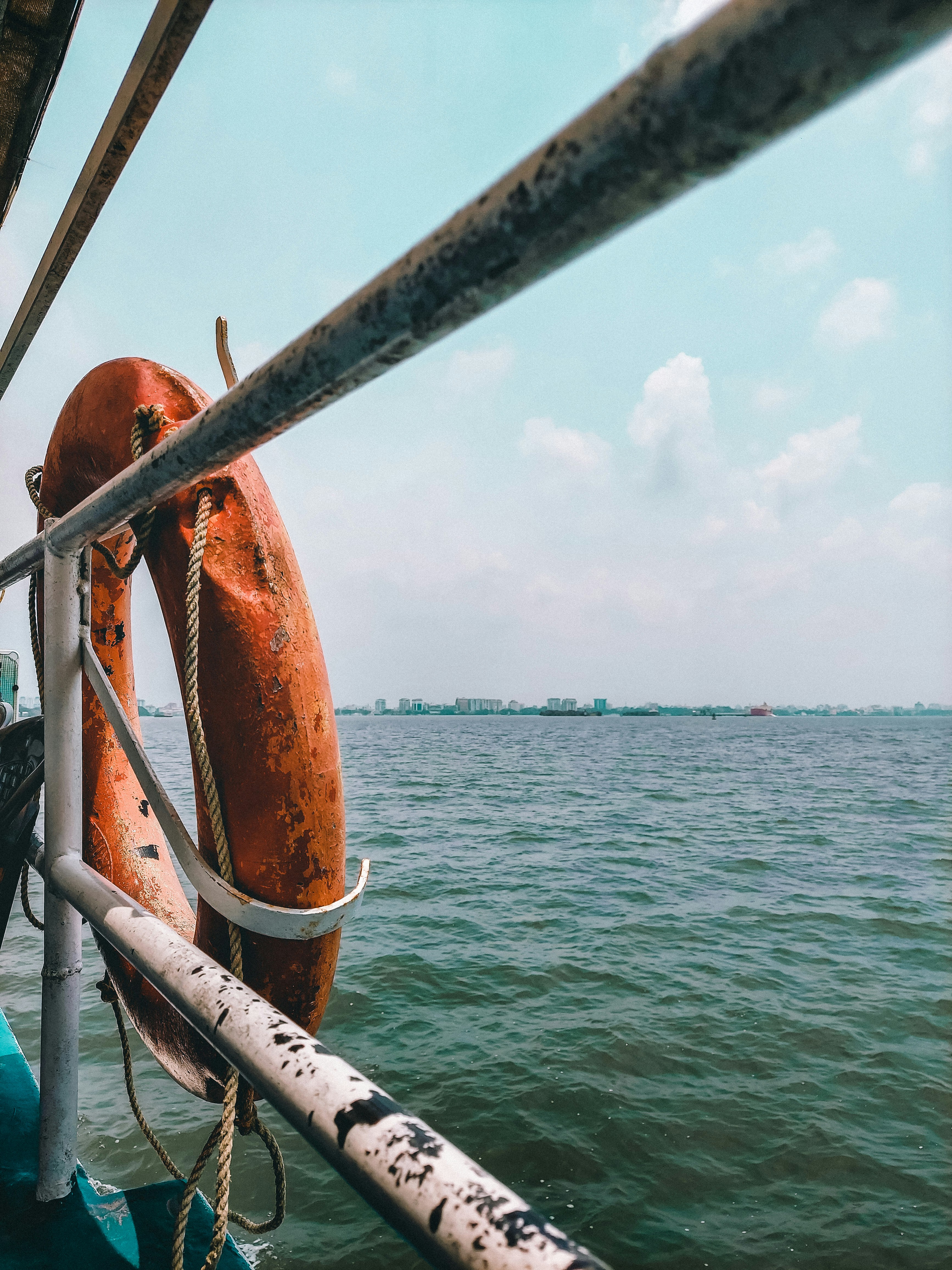 Orange lifebuoy mounted on weathered rails of a boat, with calm sea and a distant city skyline under a bright sky. The composition emphasizes nautical safety gear against a maritime horizon.