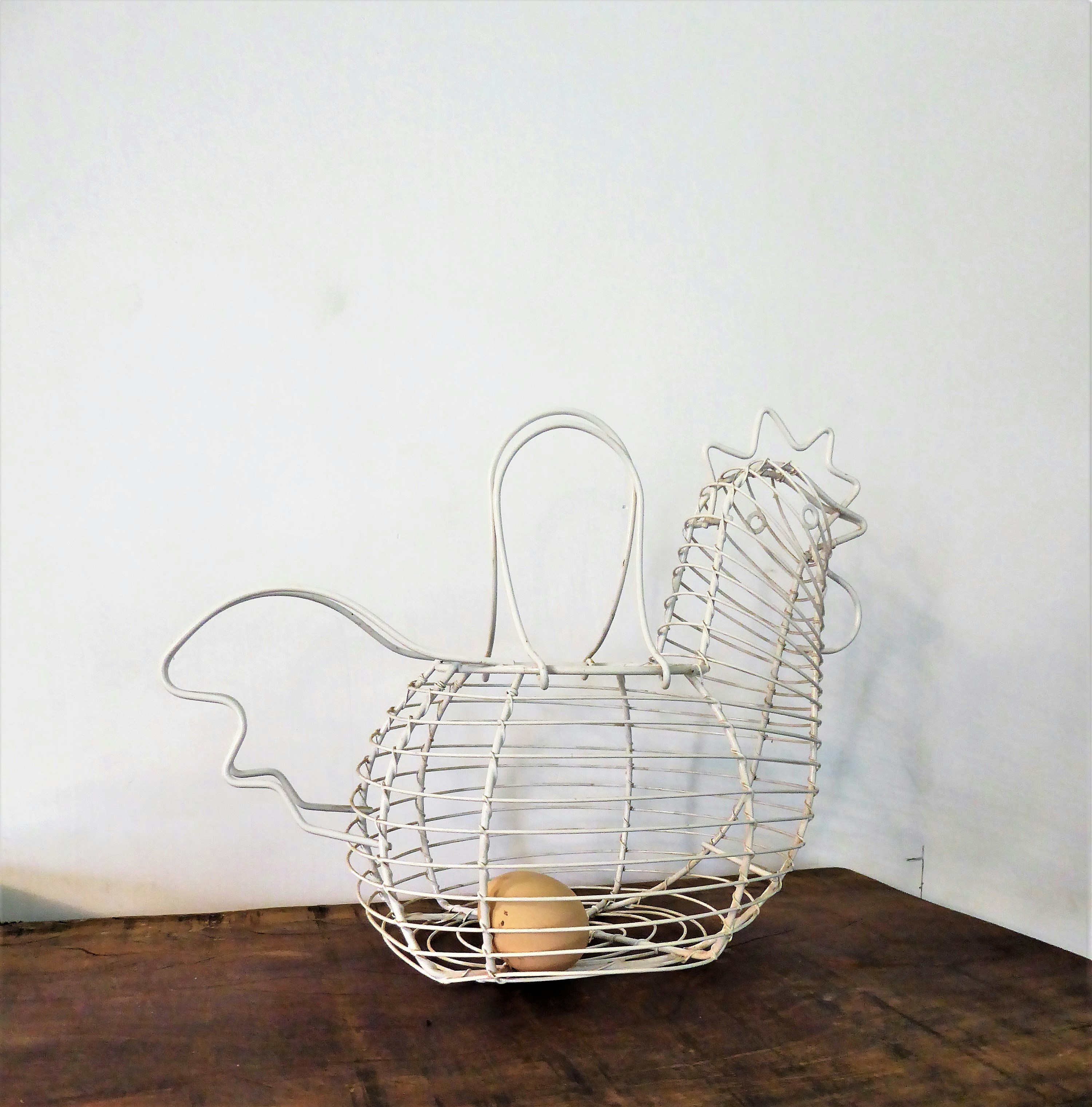 A minimalist still-life photograph of a white wire hen-shaped basket with an egg at its base, set on a dark wooden table against a pale wall.