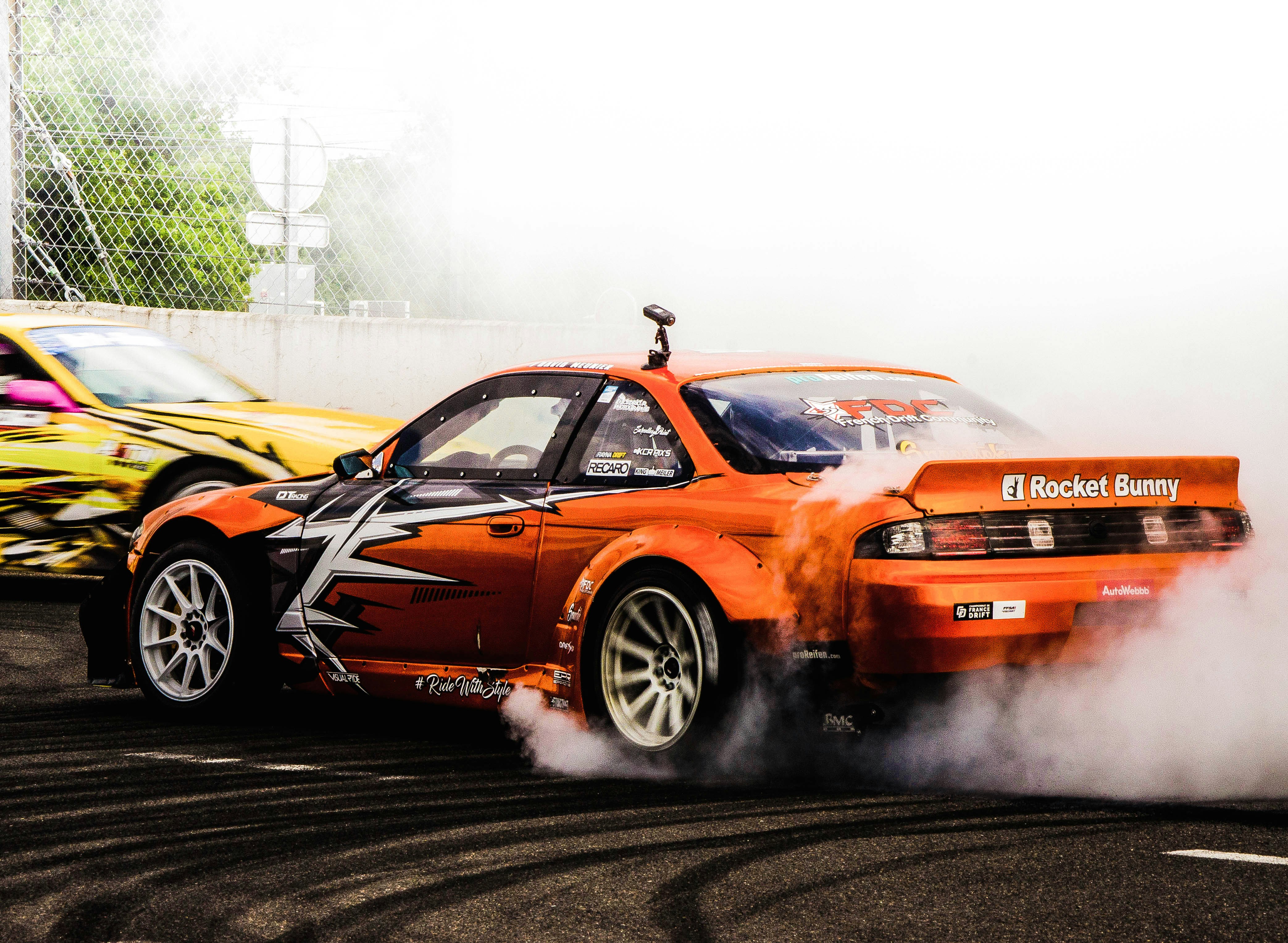 An orange drift car with Rocket Bunny branding performs a high-speed maneuver, surrounded by tire smoke on a racetrack.