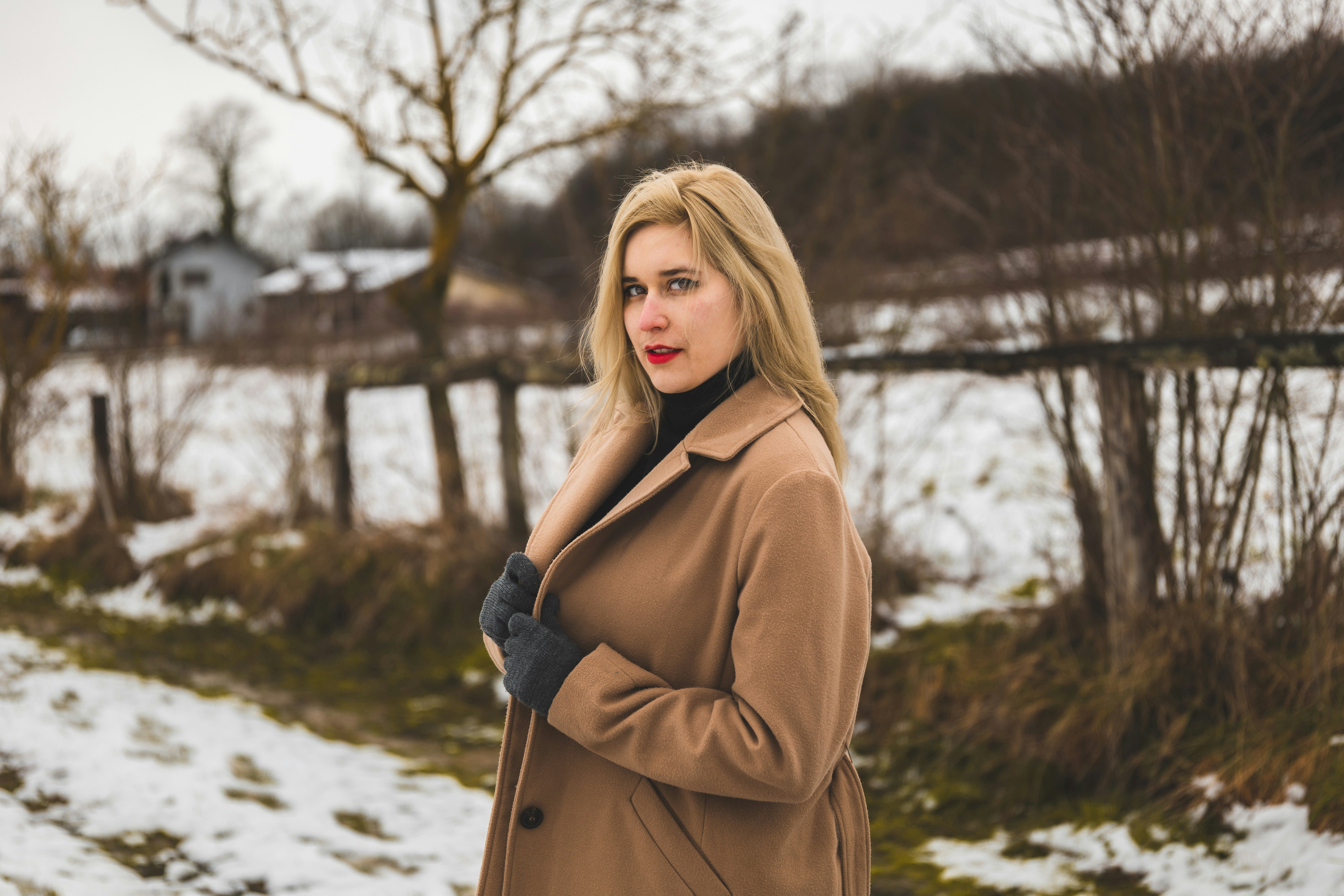 A blonde woman wearing a stylish beige coat and striking red lipstick, standing confidently in a snowy field. The winter landscape creates a beautiful and serene backdrop for this fashionable winter look. The woman exudes an air of sophistication and elegance, perfectly complementing the natural beauty of the surrounding scenery.