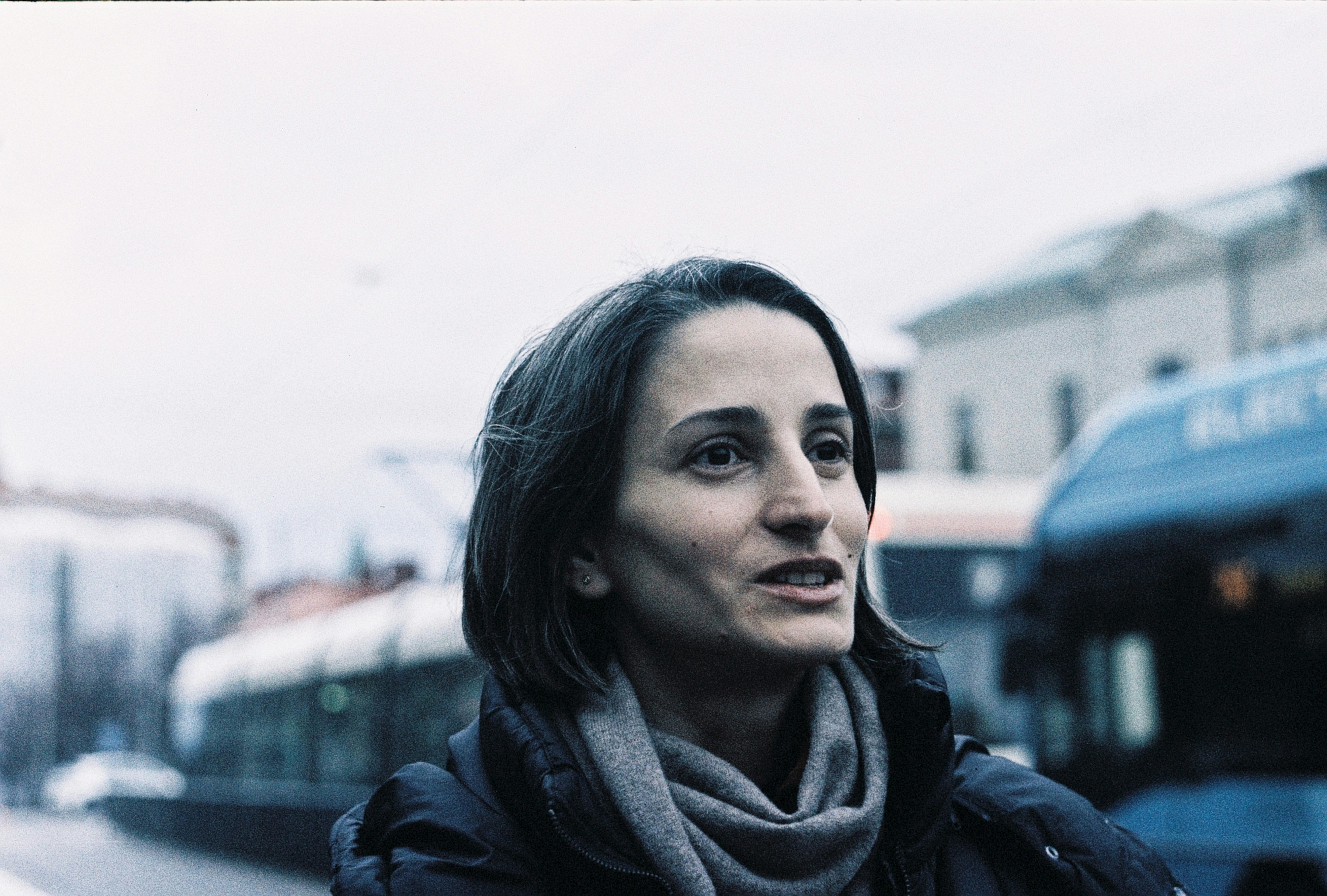 Woman with a thoughtful expression stands at a city transit stop, with blurred vehicles and buildings in the background.
