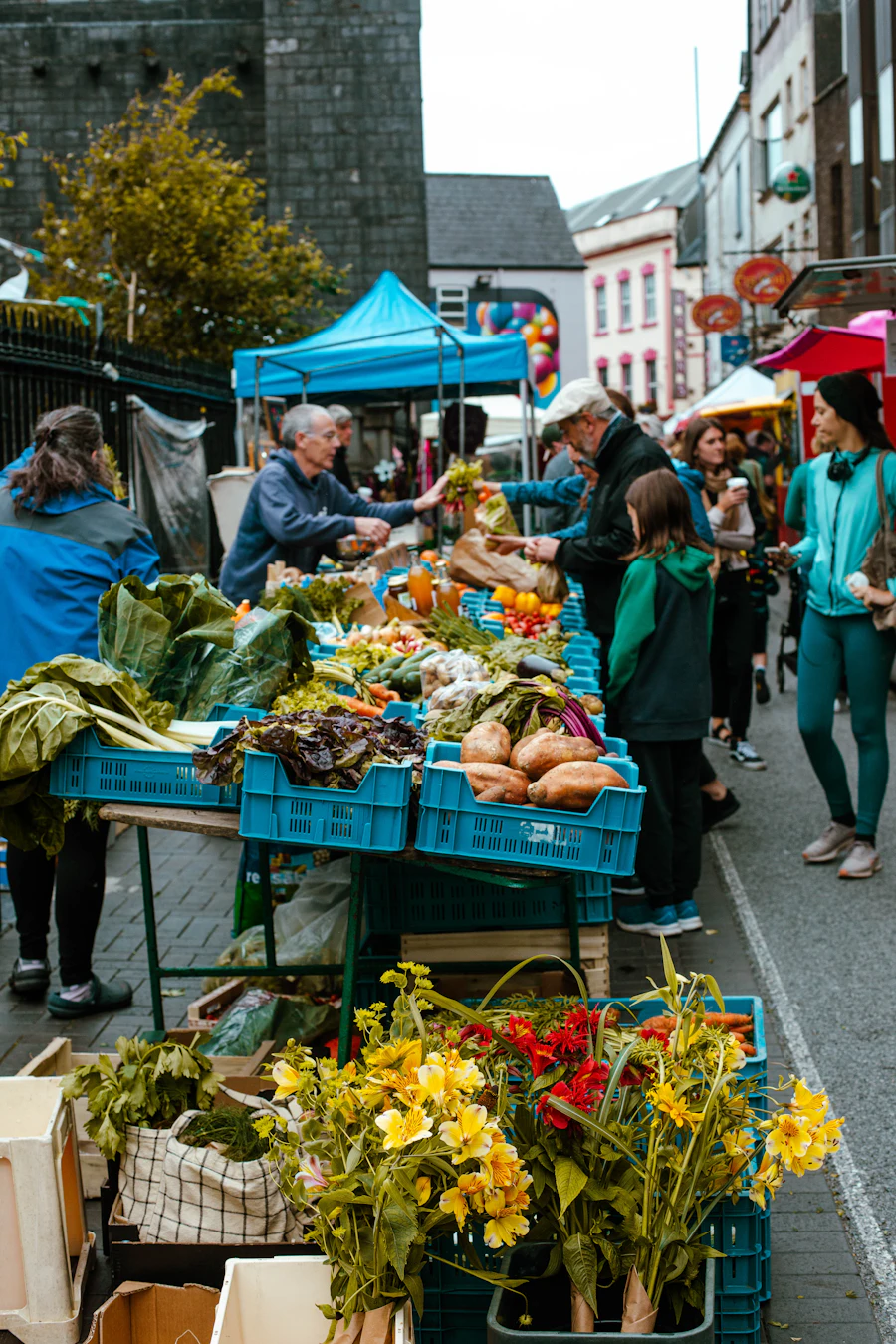 Outdoor market scene with vendors and shoppers gathered around a colorful produce stall piled with leafy greens, potatoes, and flowers. Buildings and tents line the narrow street in the background.