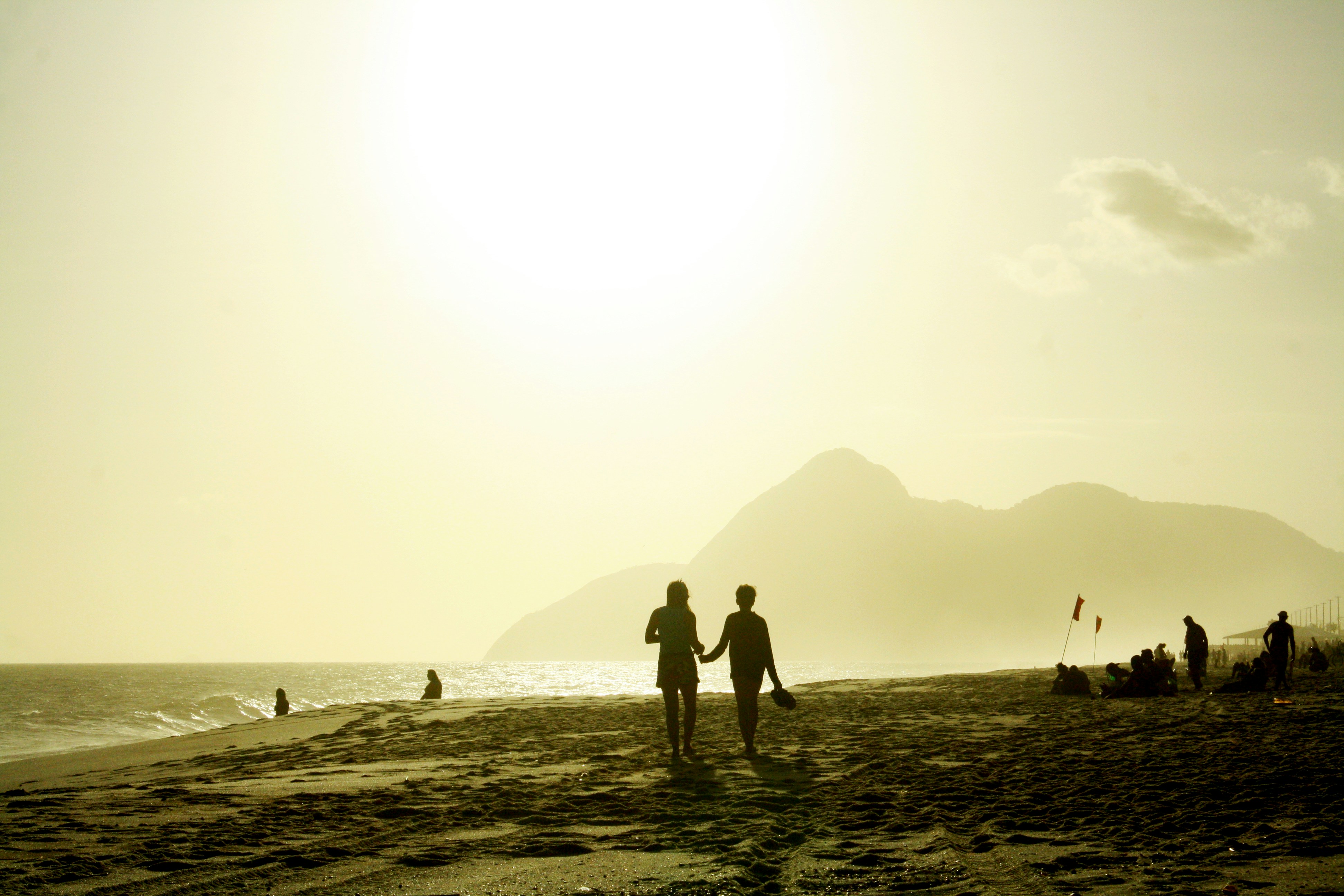 Silhouetted couple walking hand in hand on a sunlit beach with distant mountains.