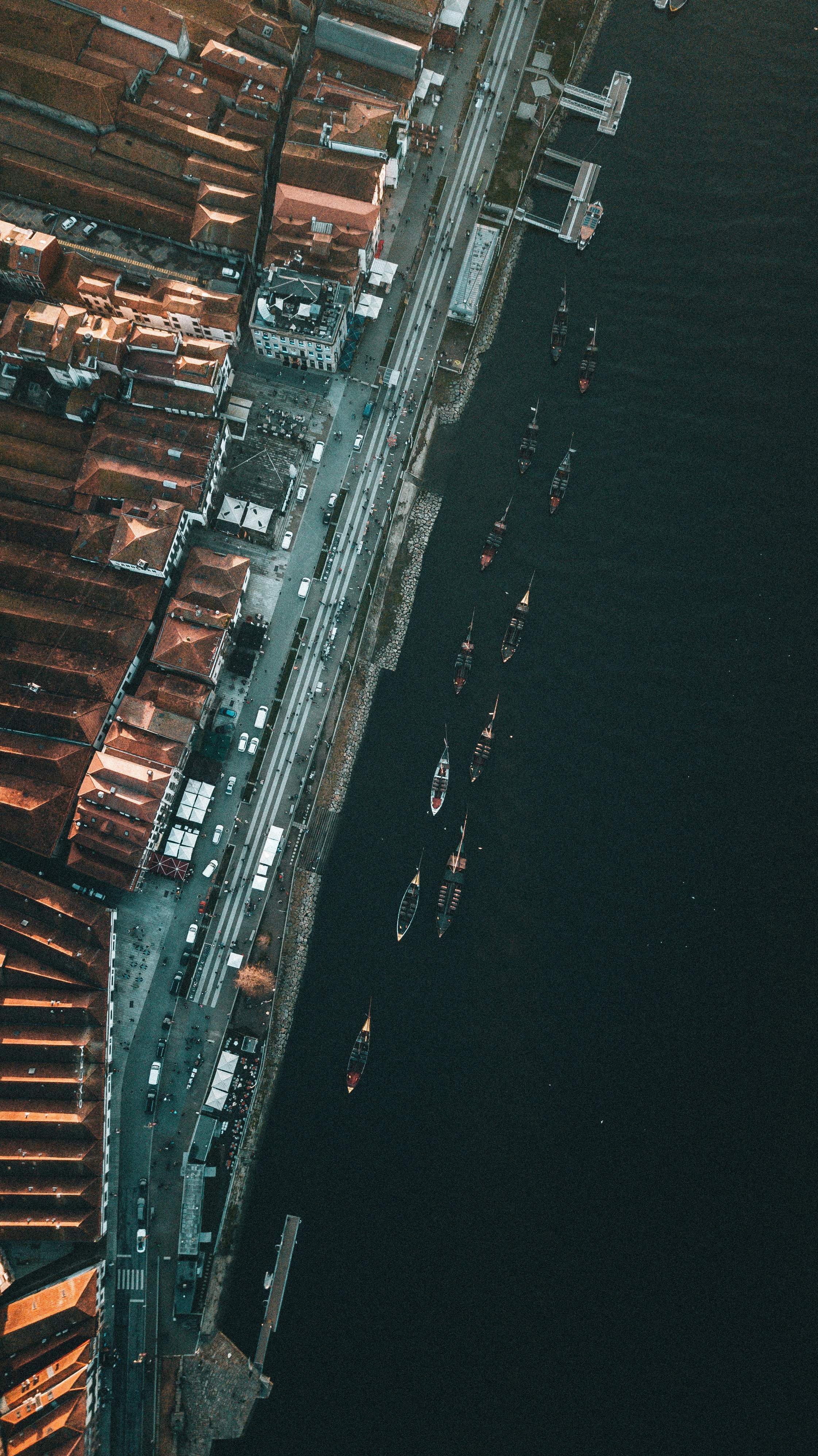 Aerial view of a row of boats moored along a city waterfront, with terracotta-roofed buildings lining the adjacent street.