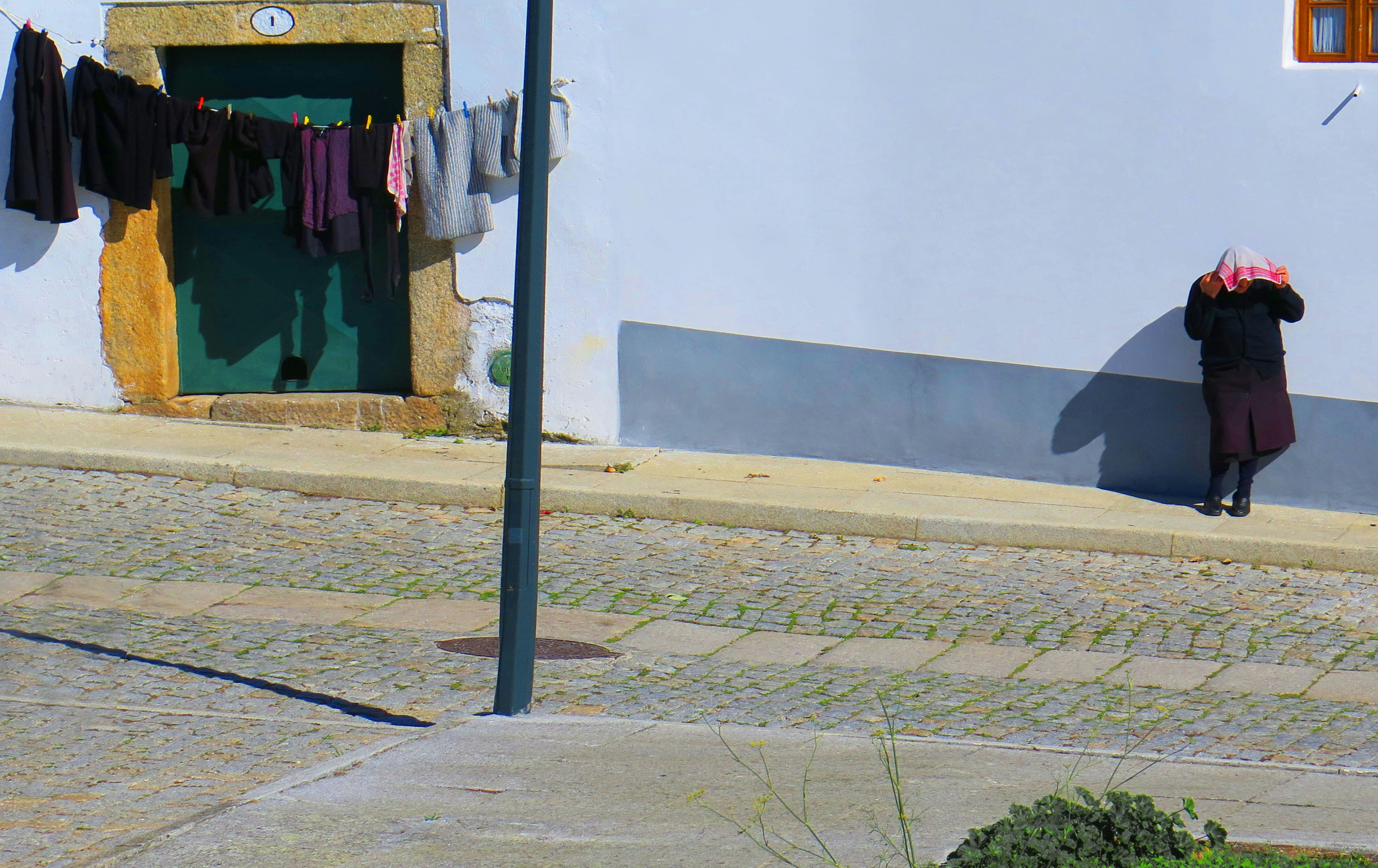 Street photograph of a sunlit blue wall with a green door, laundry hanging on a line above, and a person in a dark coat and pink hat leaning against the wall.