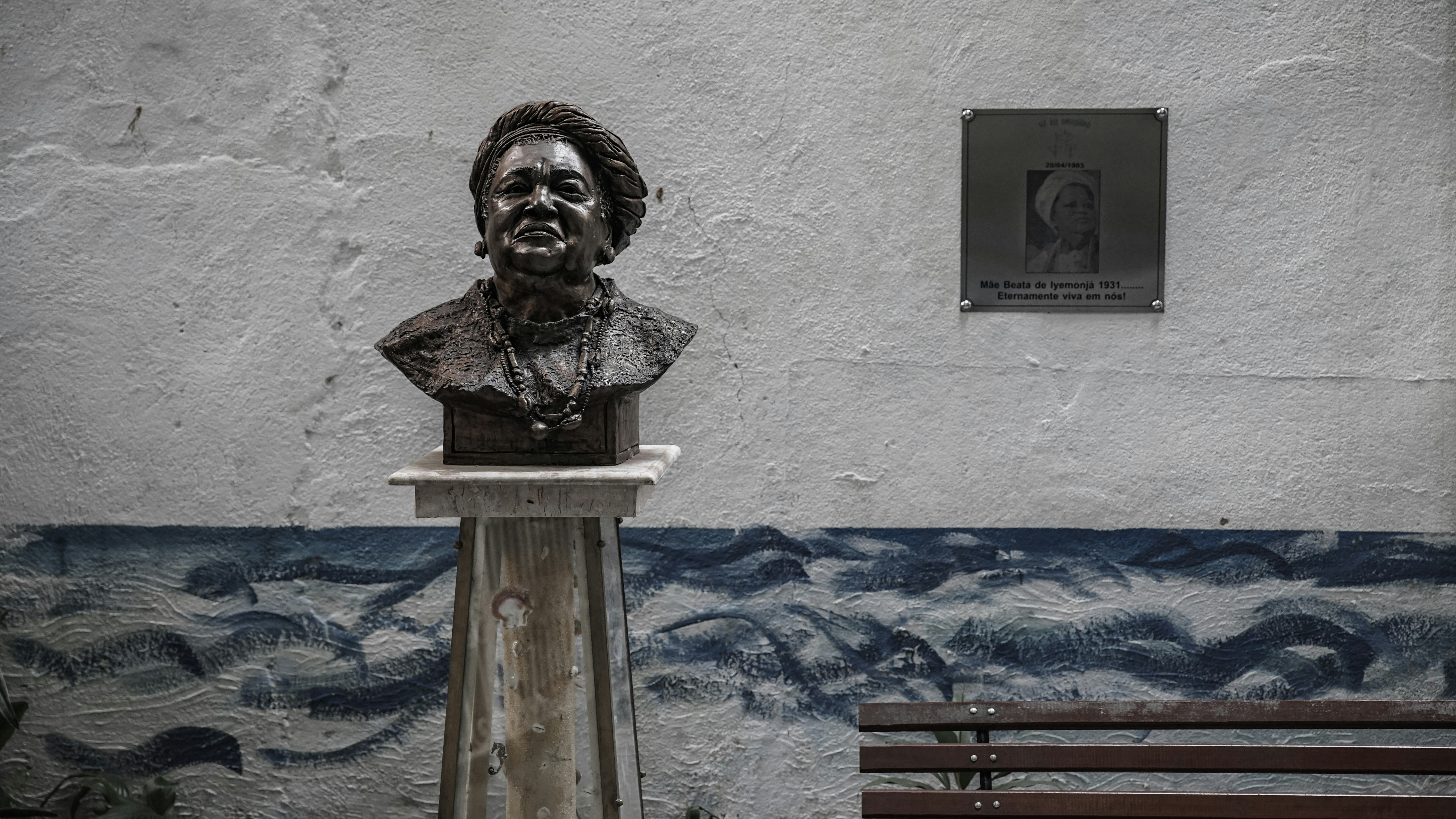 Bronze bust on a pedestal against a textured wall with a painted wave design and a wooden bench.