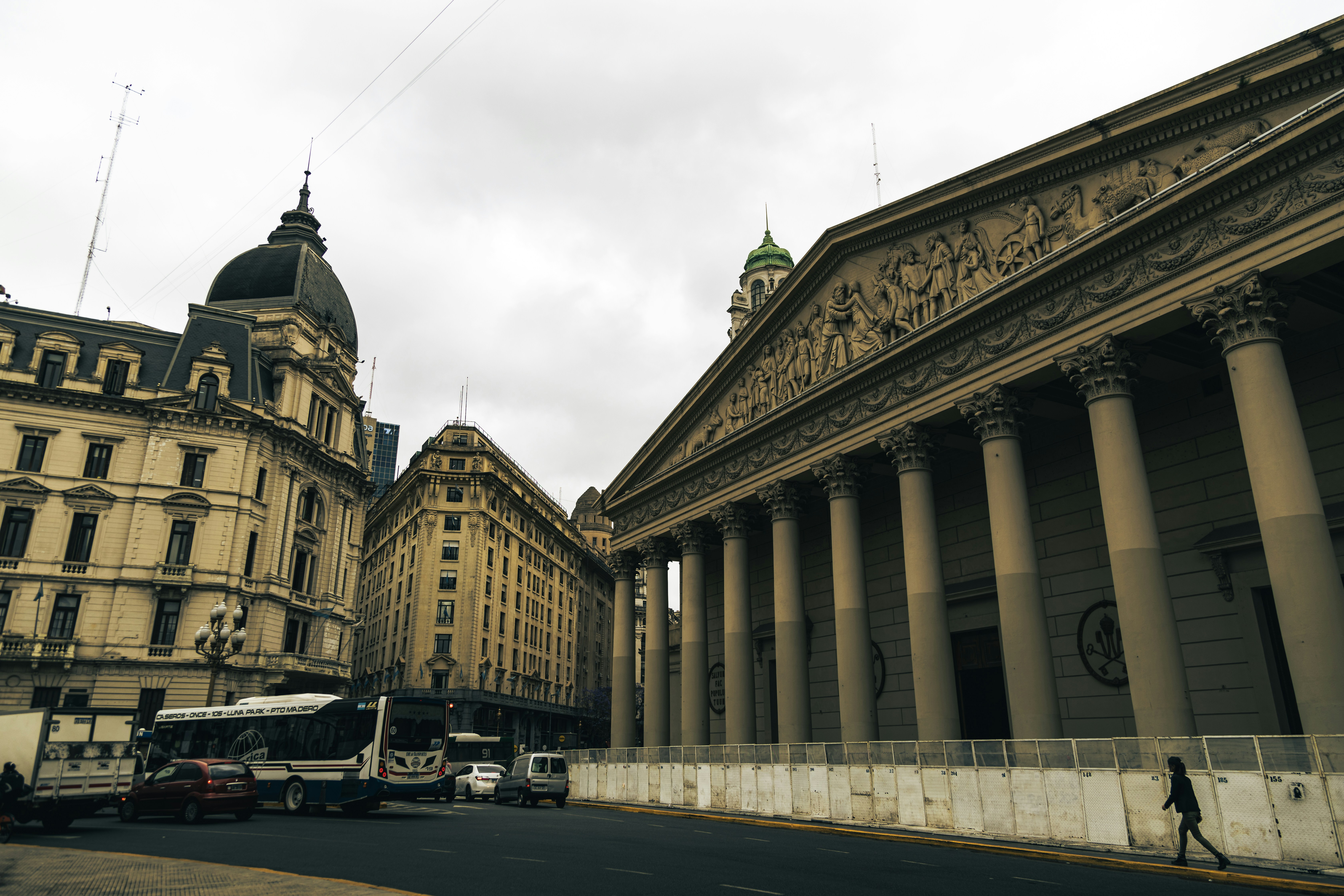 Neoclassical building with tall columns alongside historic urban architecture under a cloudy sky.