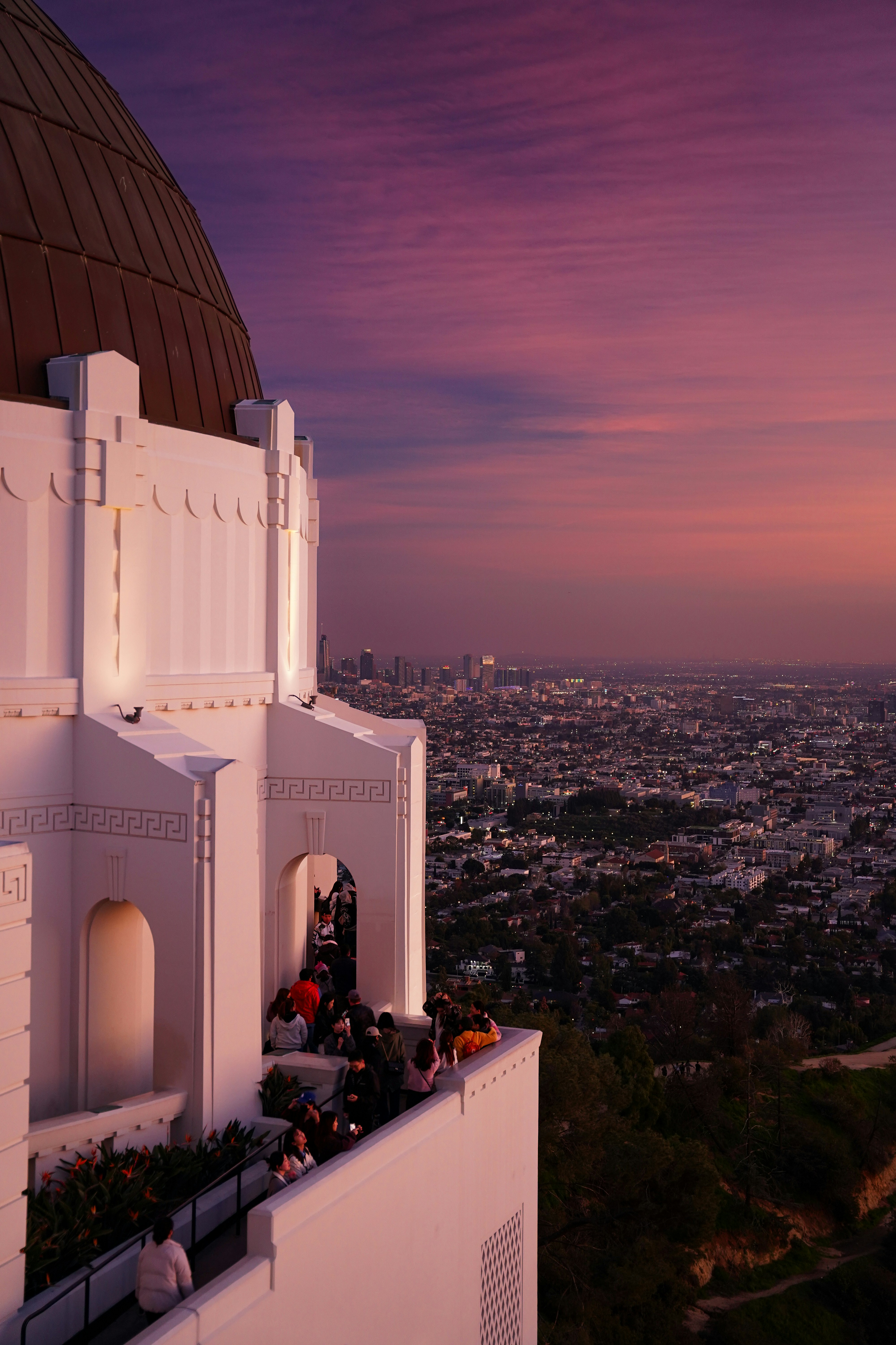 Visitors gather on the terrace of the Griffith Observatory, overlooking the sprawling city of Los Angeles at twilight. The sky is painted in hues of purple and pink.