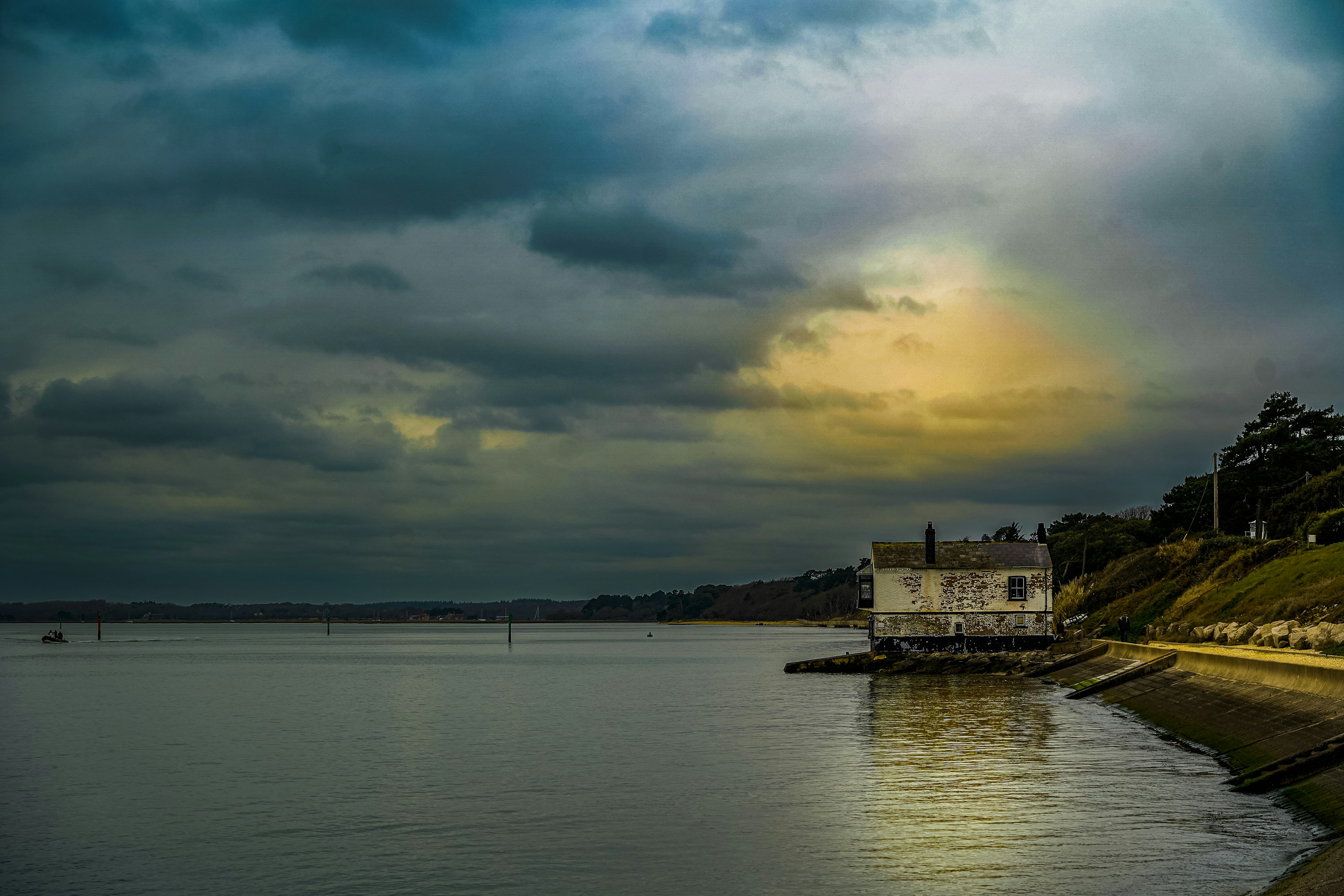 Old custom house by the calm shoreline under a dramatic, cloud-filled sky at sunset.