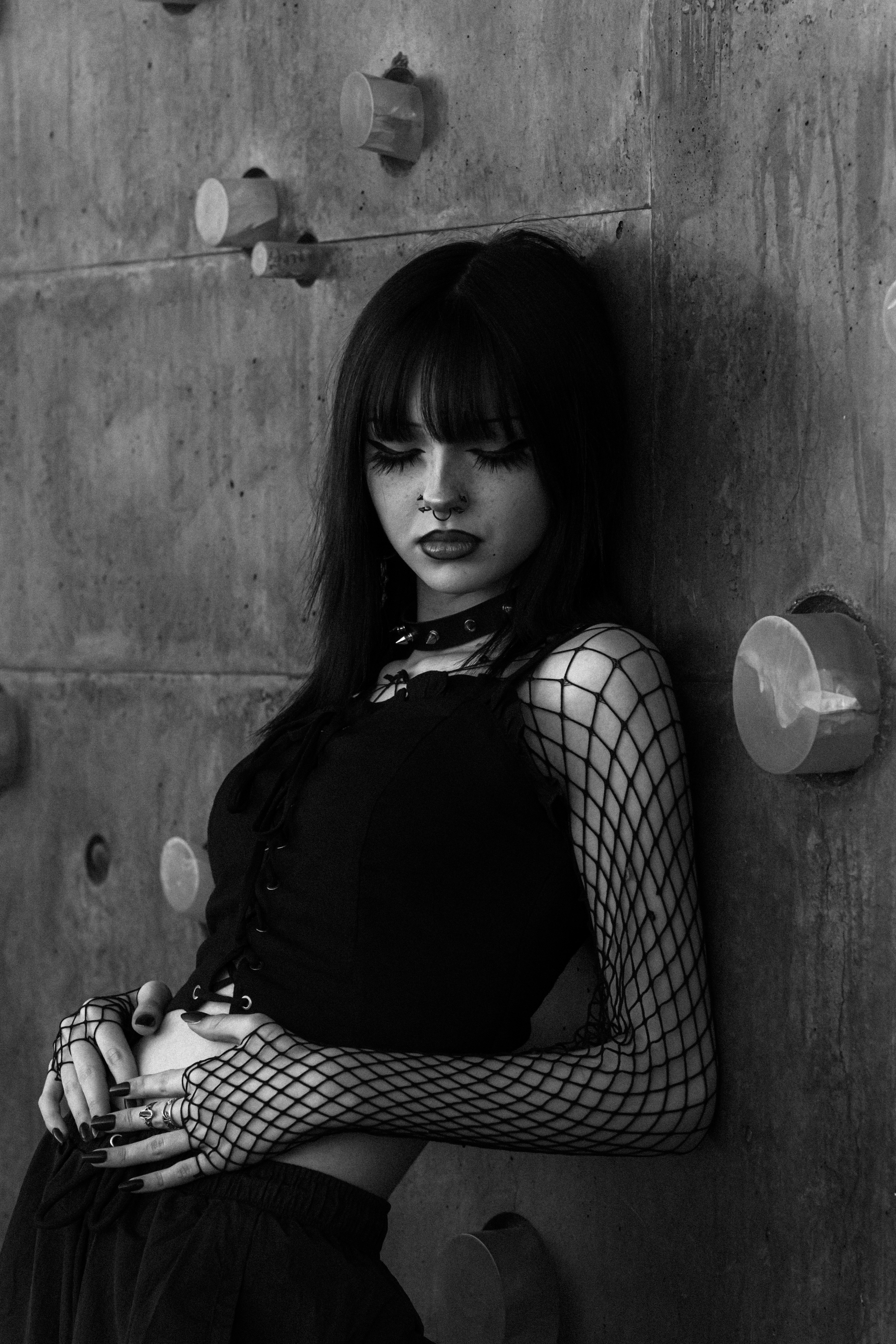 Model in a black outfit with fishnet sleeves leans against a textured concrete wall, exuding a moody aesthetic. The monochrome palette enhances the striking contrast of her look.