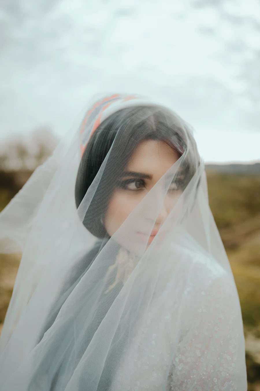 A woman outdoors is draped in a sheer veil, with her face partially hidden and eyes turned to the side. Soft natural light and a blurred landscape create a dreamy, muted mood.