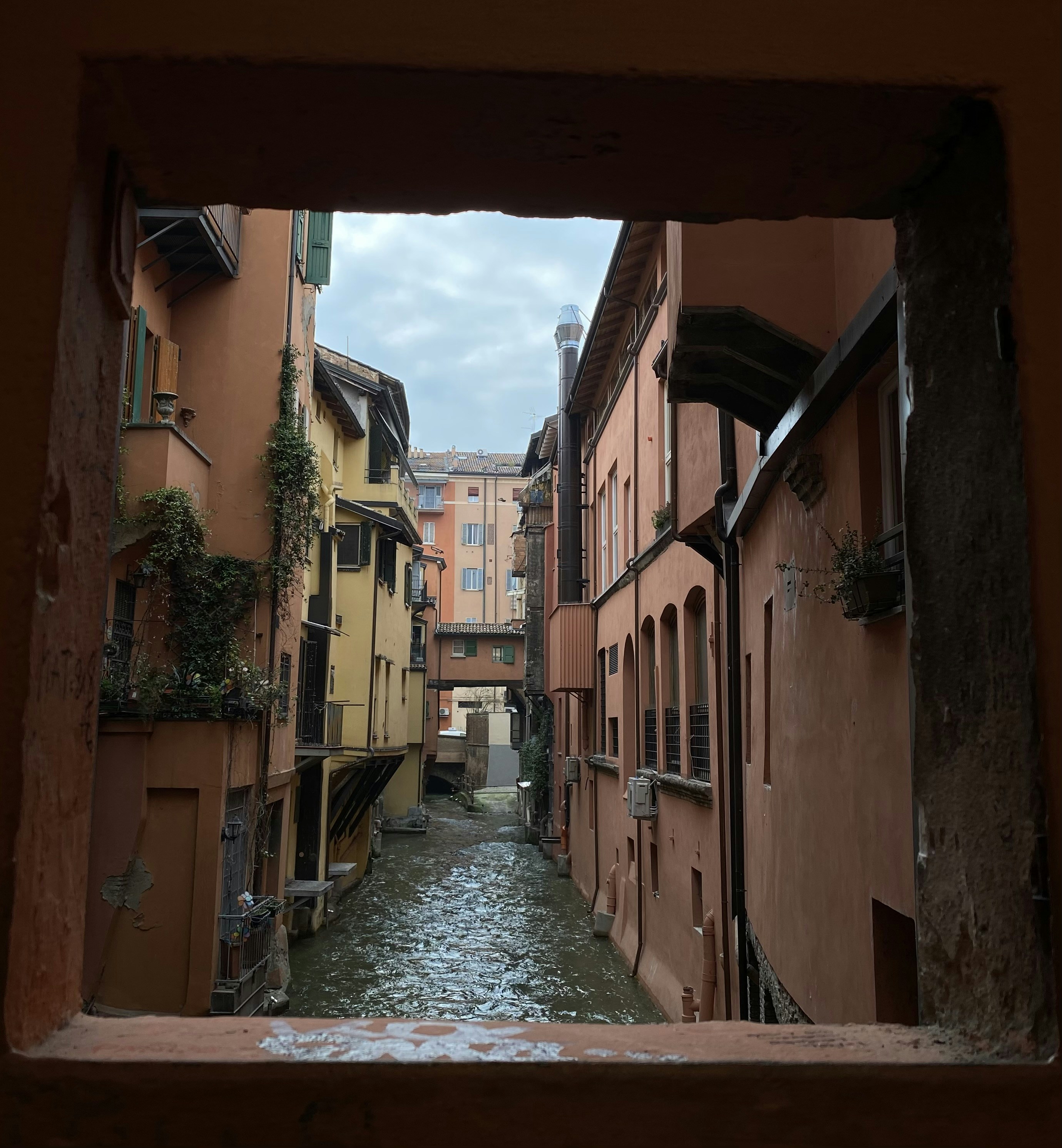 View through a stone-framed opening, showcasing colorful buildings lining a serene waterway in Bologna. 
