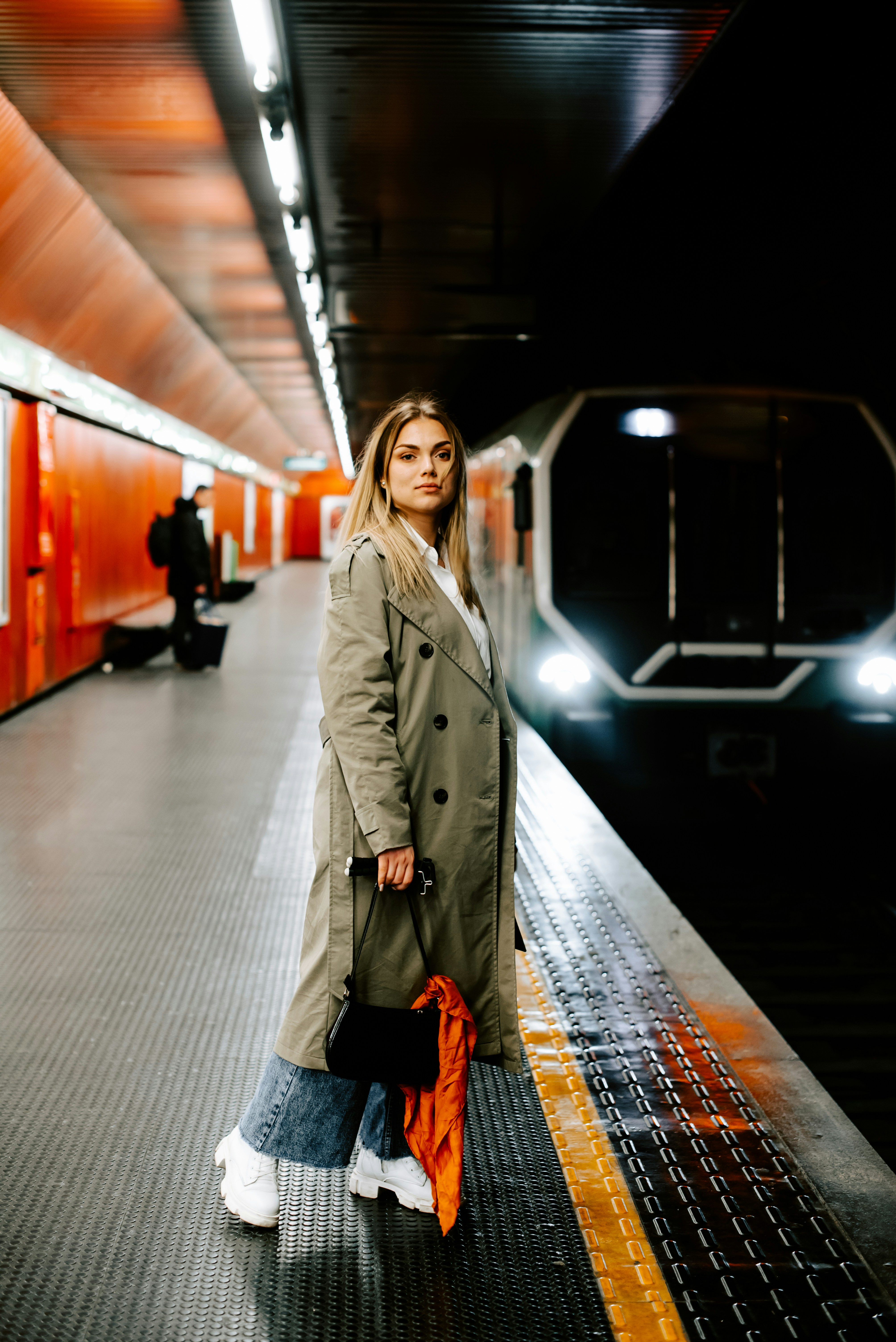 Platform Quiet: A Woman in a Trench Coat Stands Between Train Lights