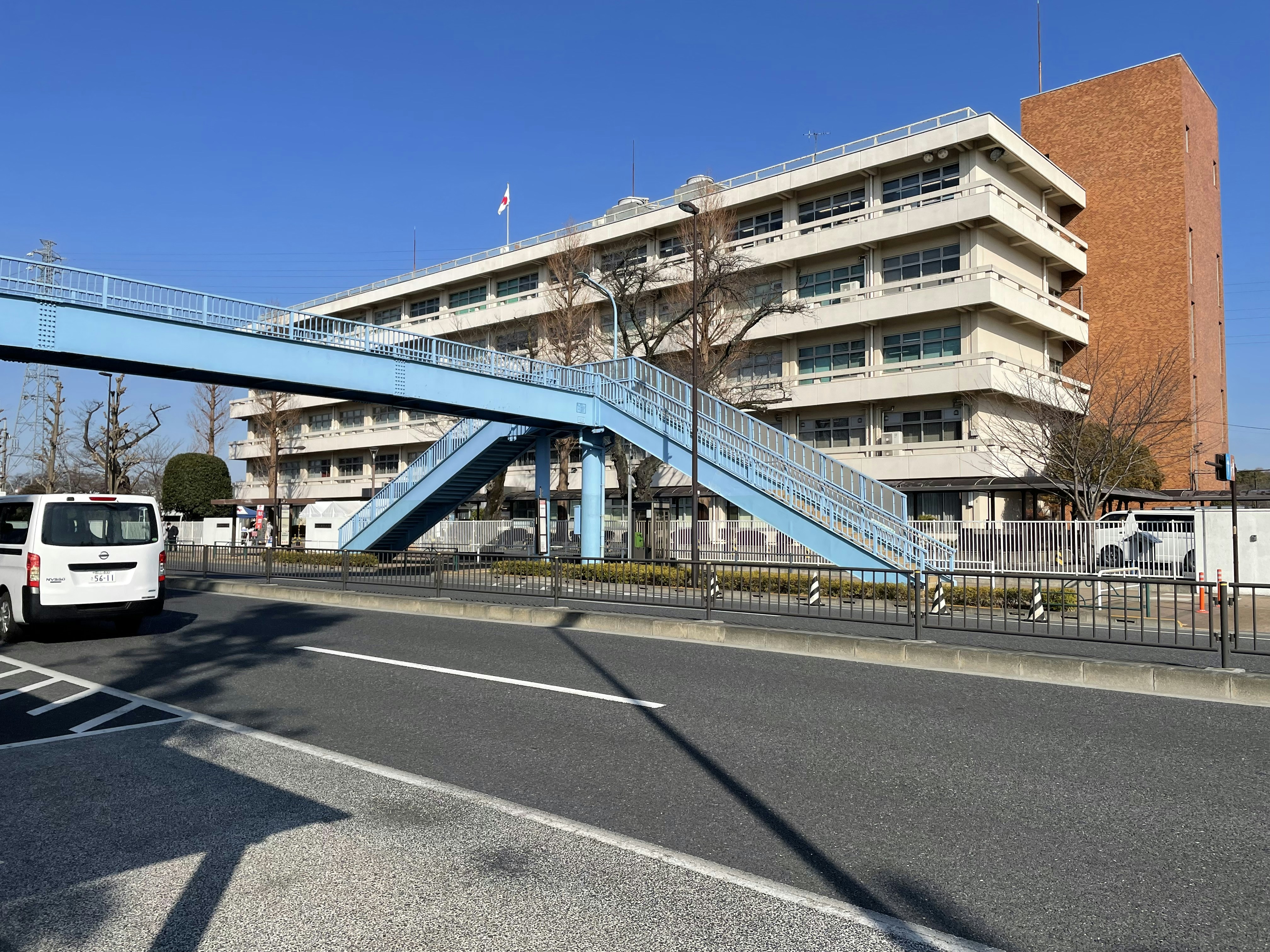 White van driving beneath a blue pedestrian bridge beside a modern building on a sunny day.