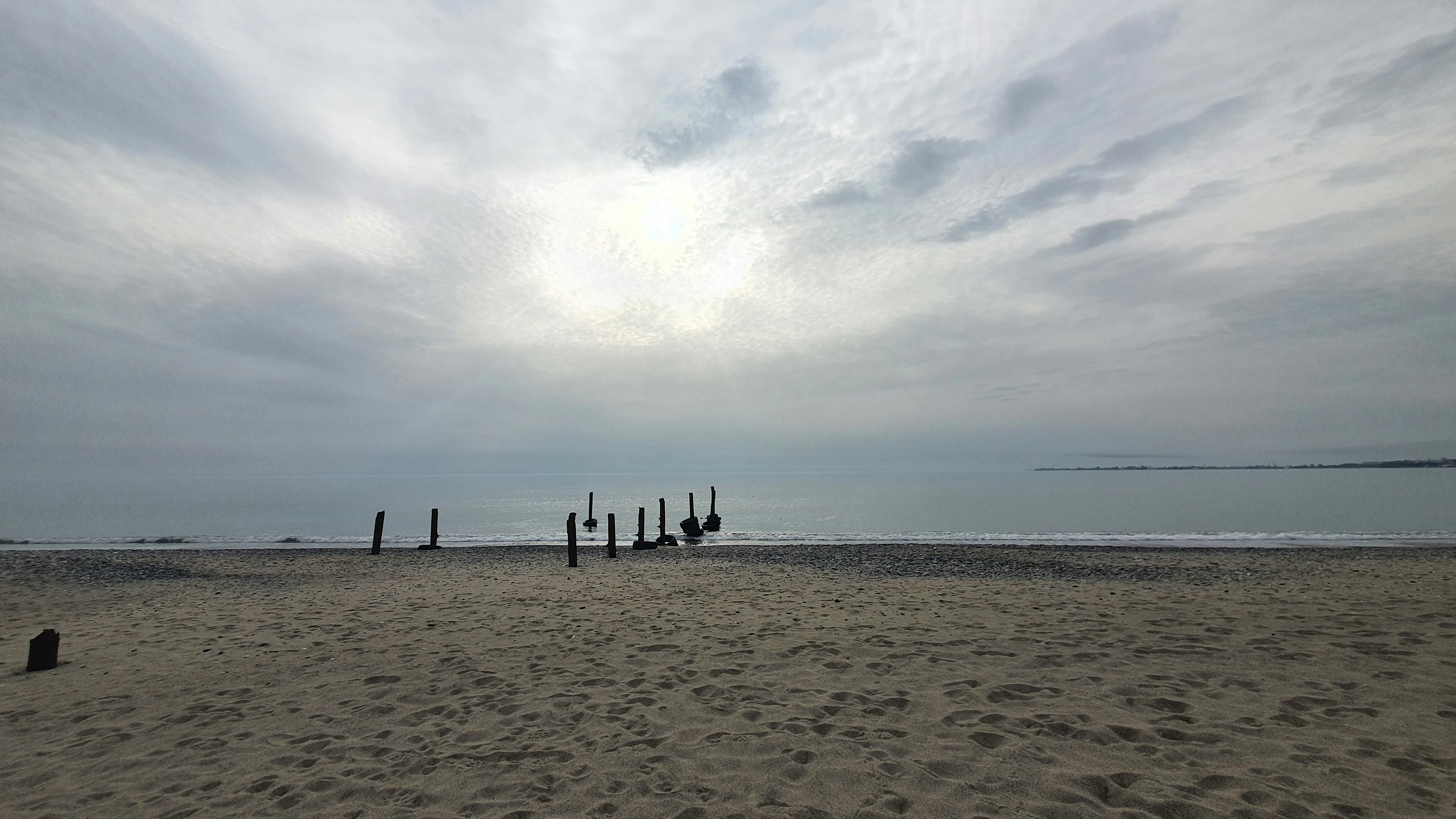 Beige-sand beach with weathered pilings extending into a calm sea beneath a muted, cloudy sky.