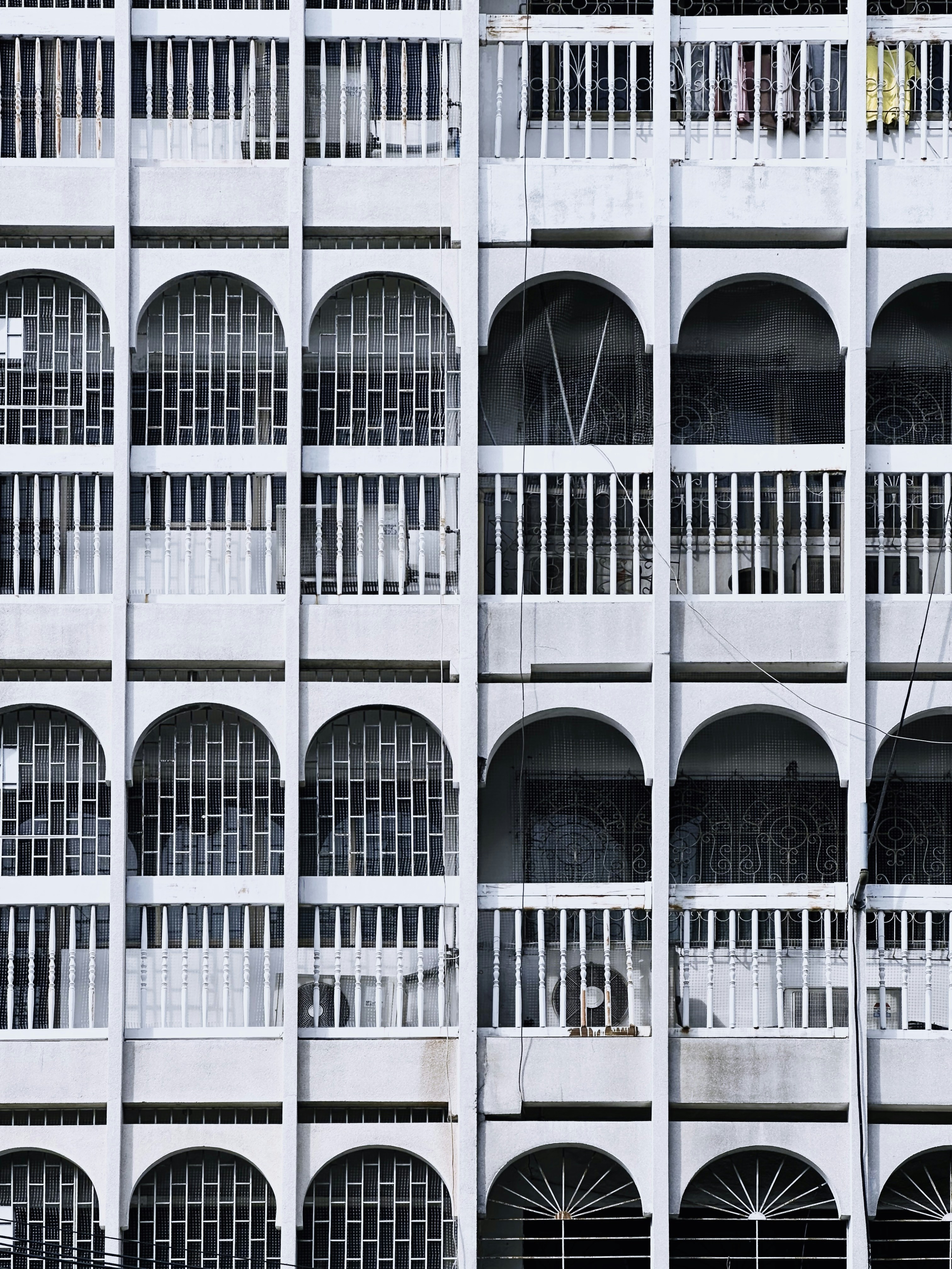 Geometric facade of a multi-story building with repeating arched windows and metal grilles, forming a grid-like pattern across the surface. Aerial or street-level view emphasizes the architectural rhythm with subtle weathering.