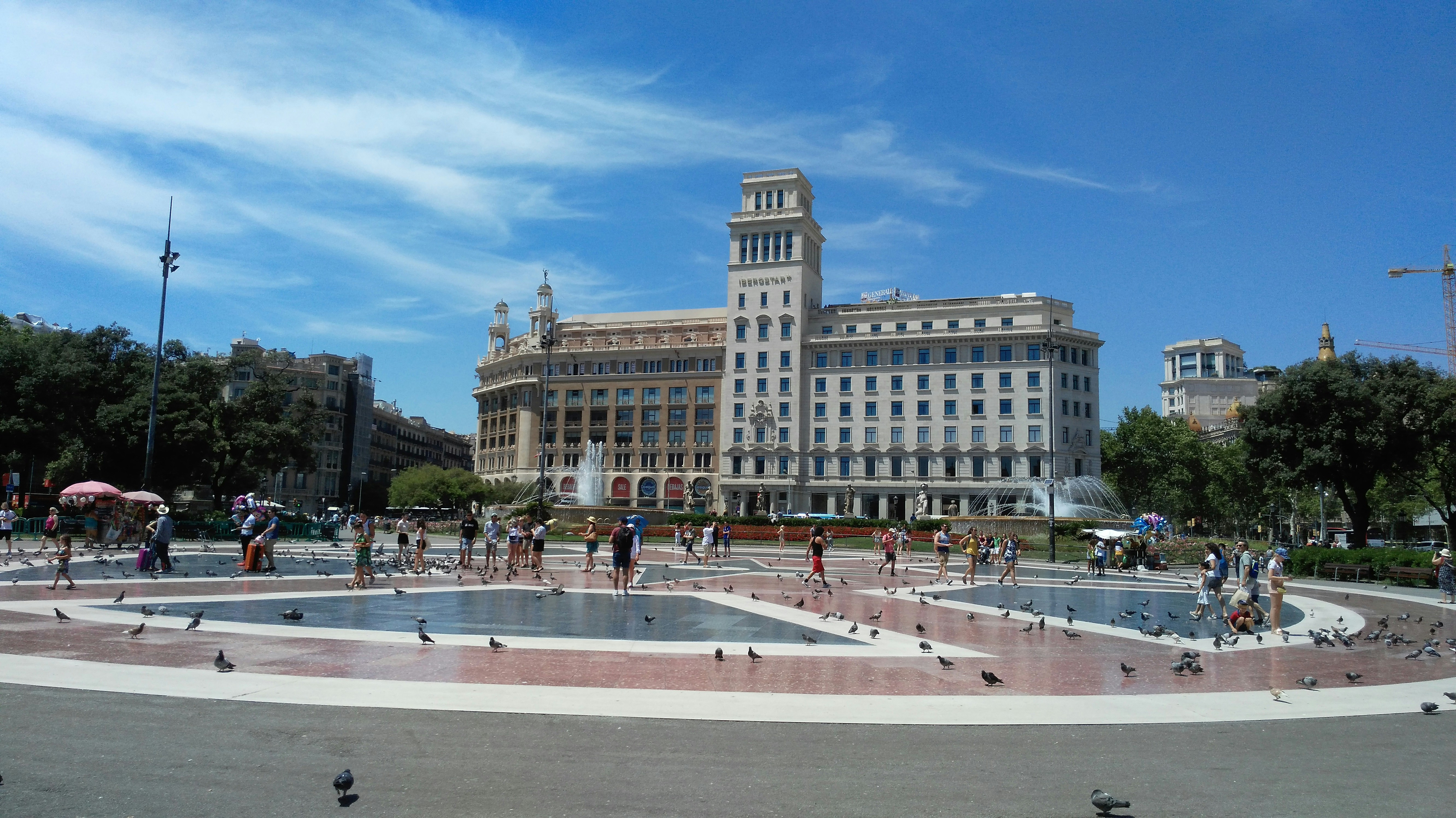 A sunlit city square features a circular fountain in the foreground and a tall, towered building rising against a clear blue sky, with people strolling across the plaza.