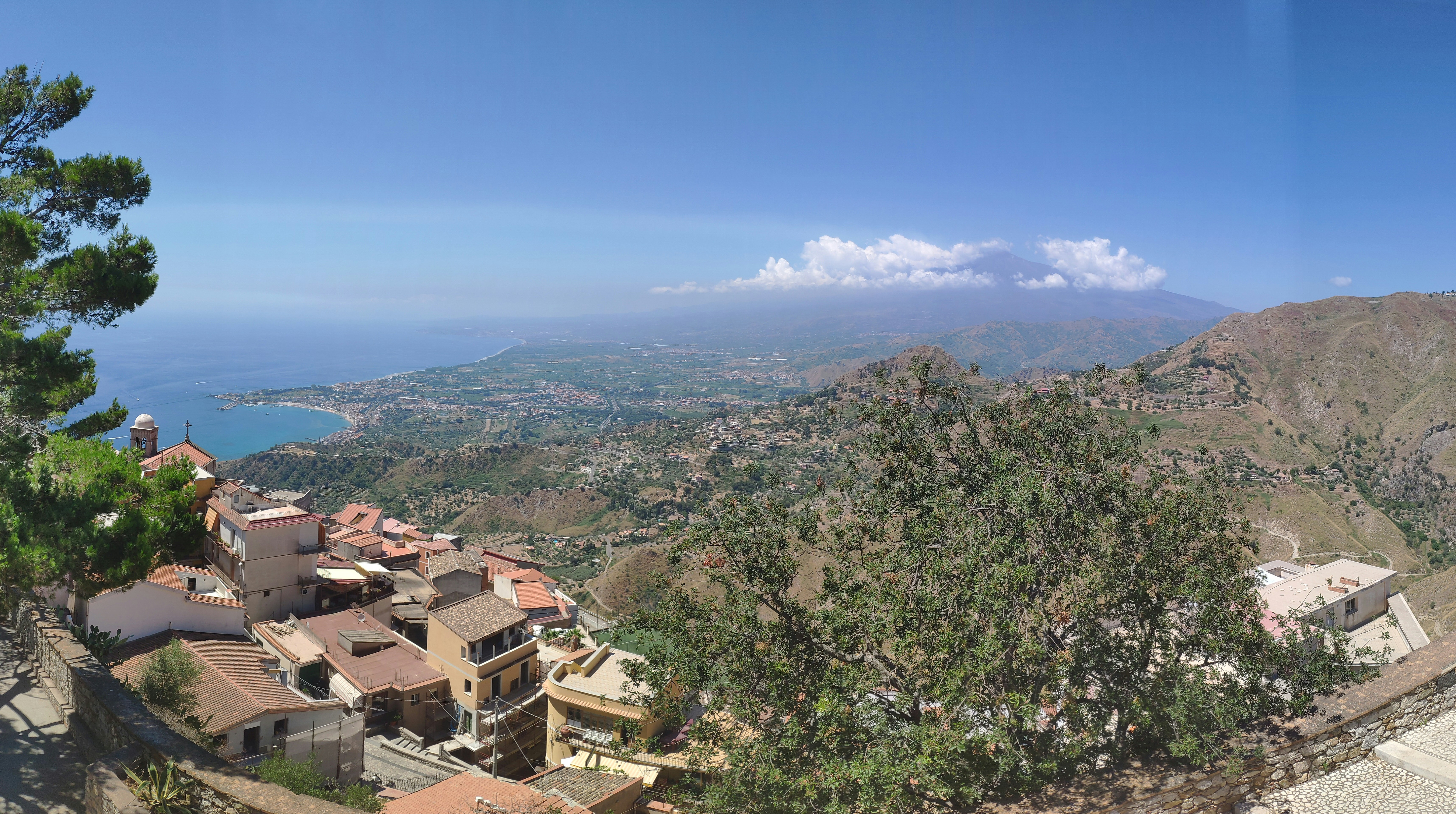 Panoramic view of a coastal town in Sicily, showcasing a blend of terracotta rooftops and lush greenery against a backdrop of the Mediterranean Sea and distant mountains.