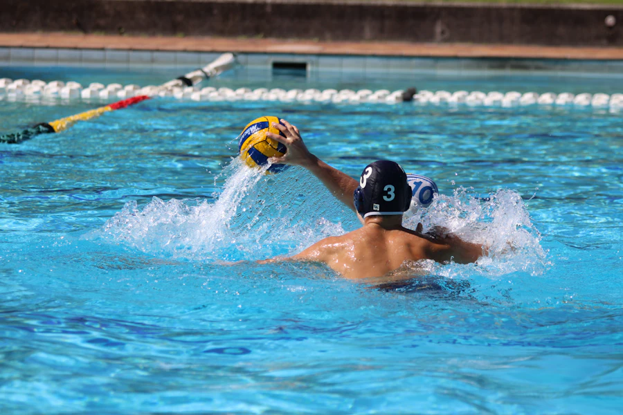 Water polo player wearing cap number 3 reaching to throw a yellow ball over splashing pool water in bright daylight.
