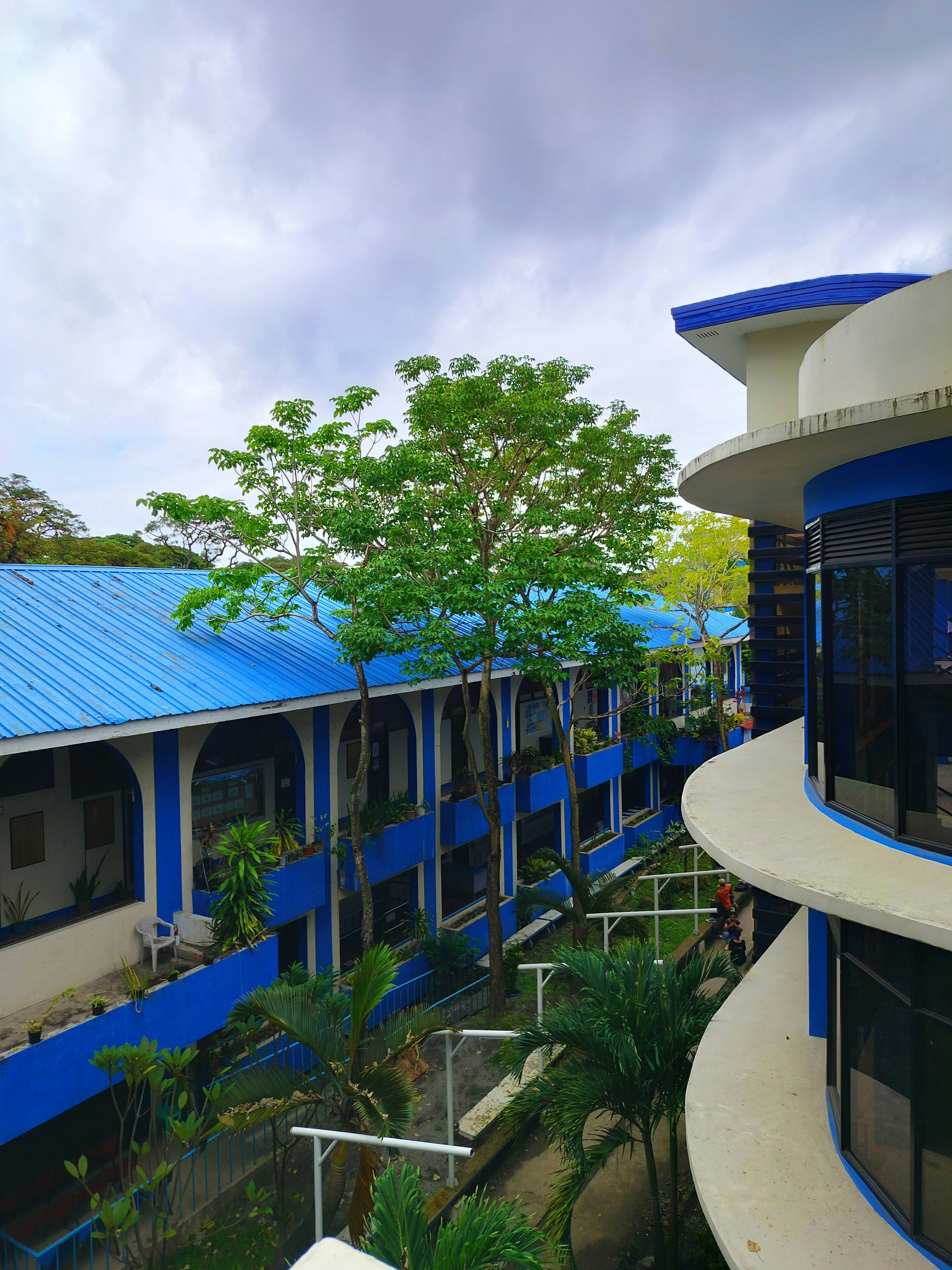 Elevated view of a blue-roofed campus building with curved white balconies and a central tree-lined courtyard.