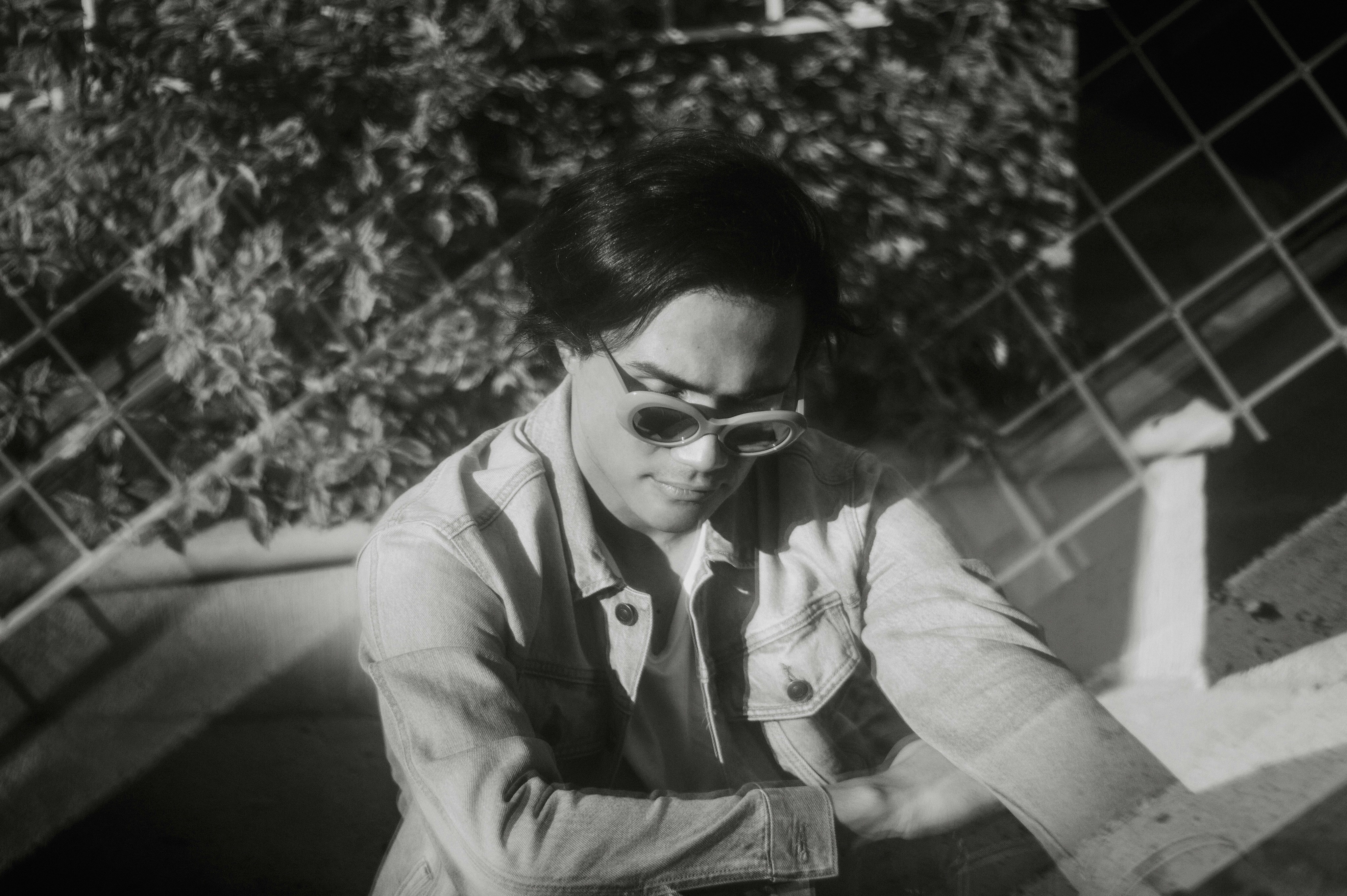 A young man in sunglasses sits pensively, surrounded by a lush backdrop of greenery and geometric patterns. The monochrome tones emphasize a moment of introspection.