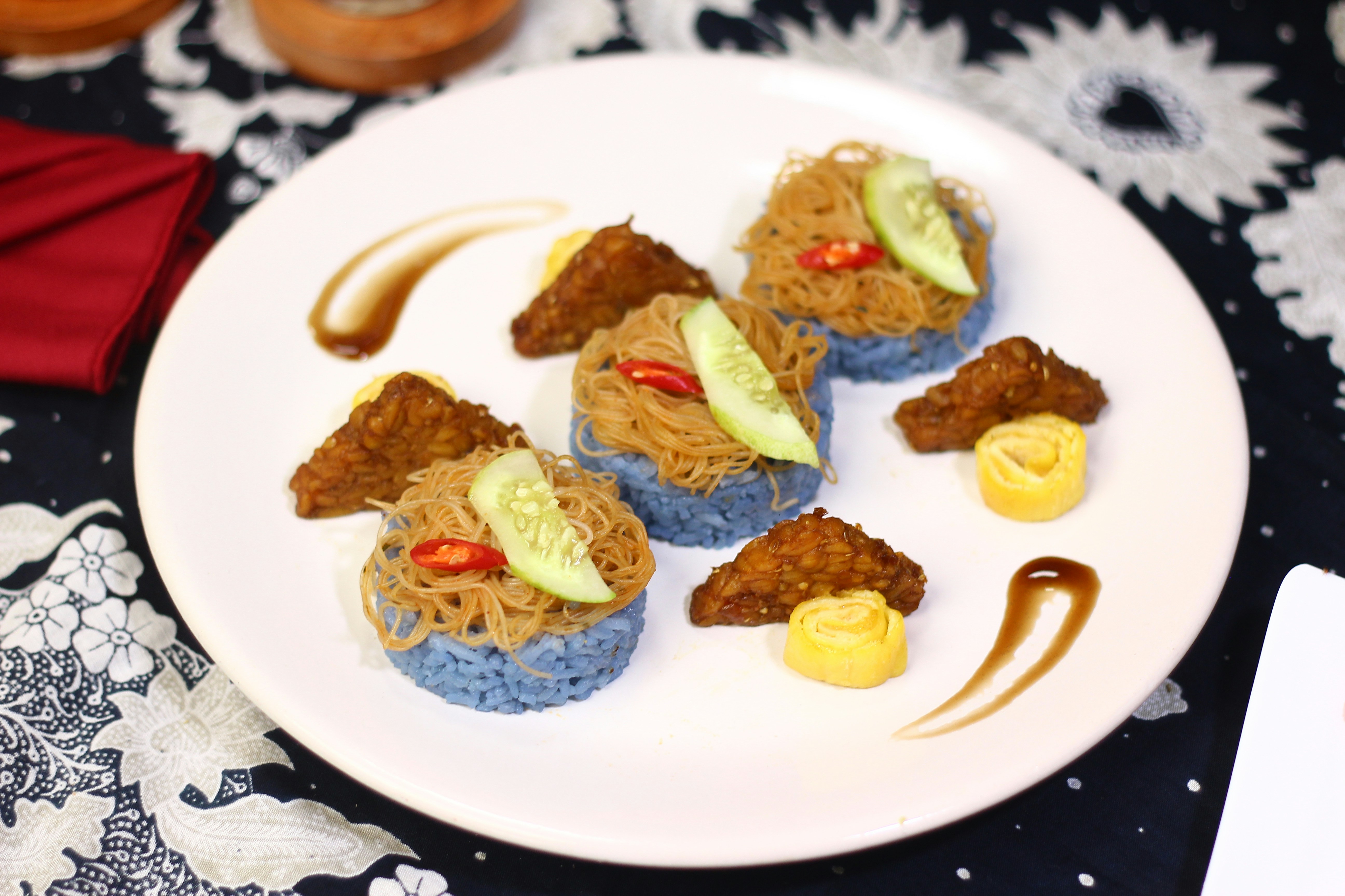 A beautifully arranged plate featuring blue rice topped with noodles, fried tempeh, and garnished with cucumber slices and chili peppers.