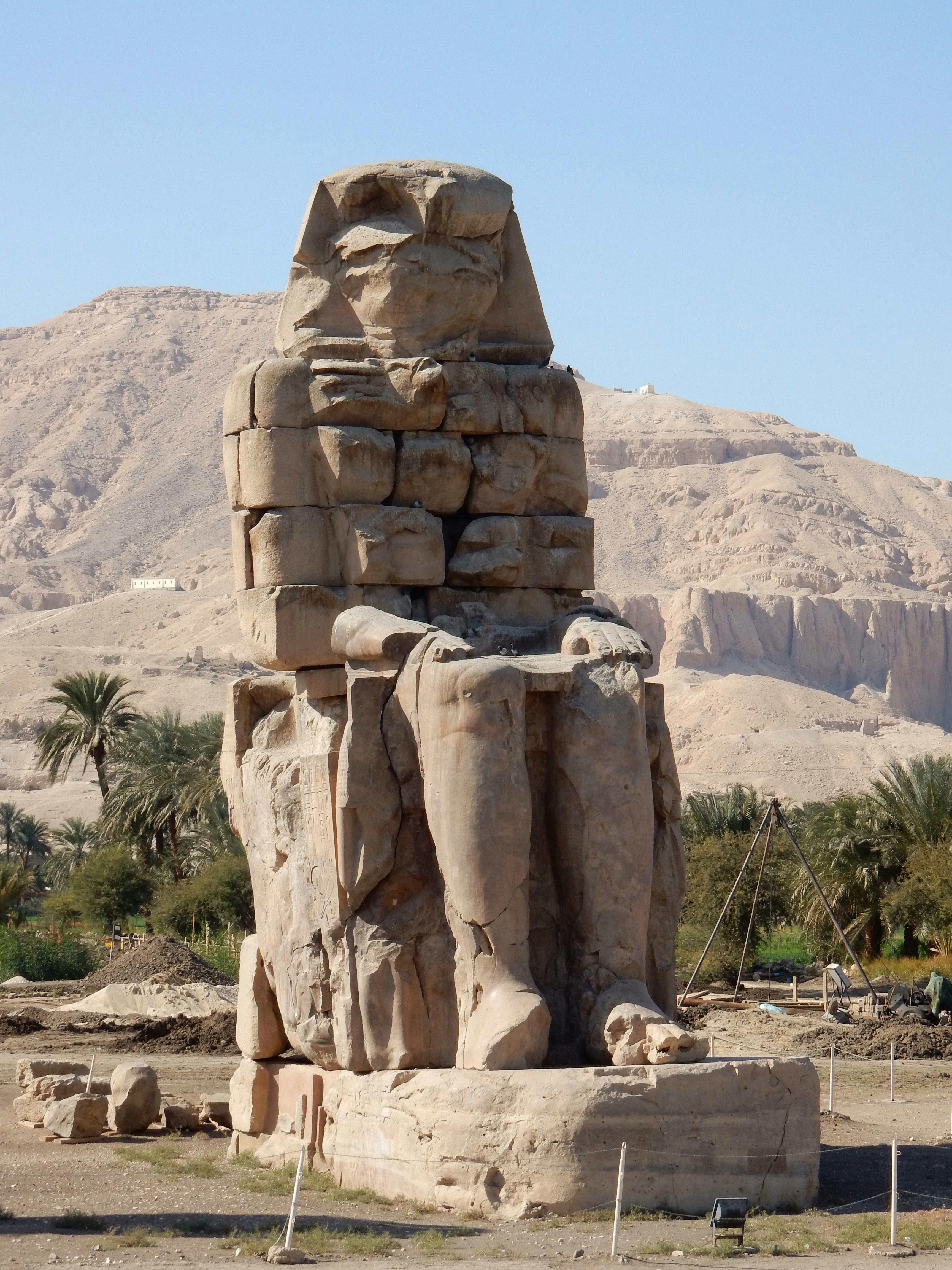 Massive ancient statue seated amidst a desert landscape, showcasing intricate stonework against a backdrop of mountains and palm trees.