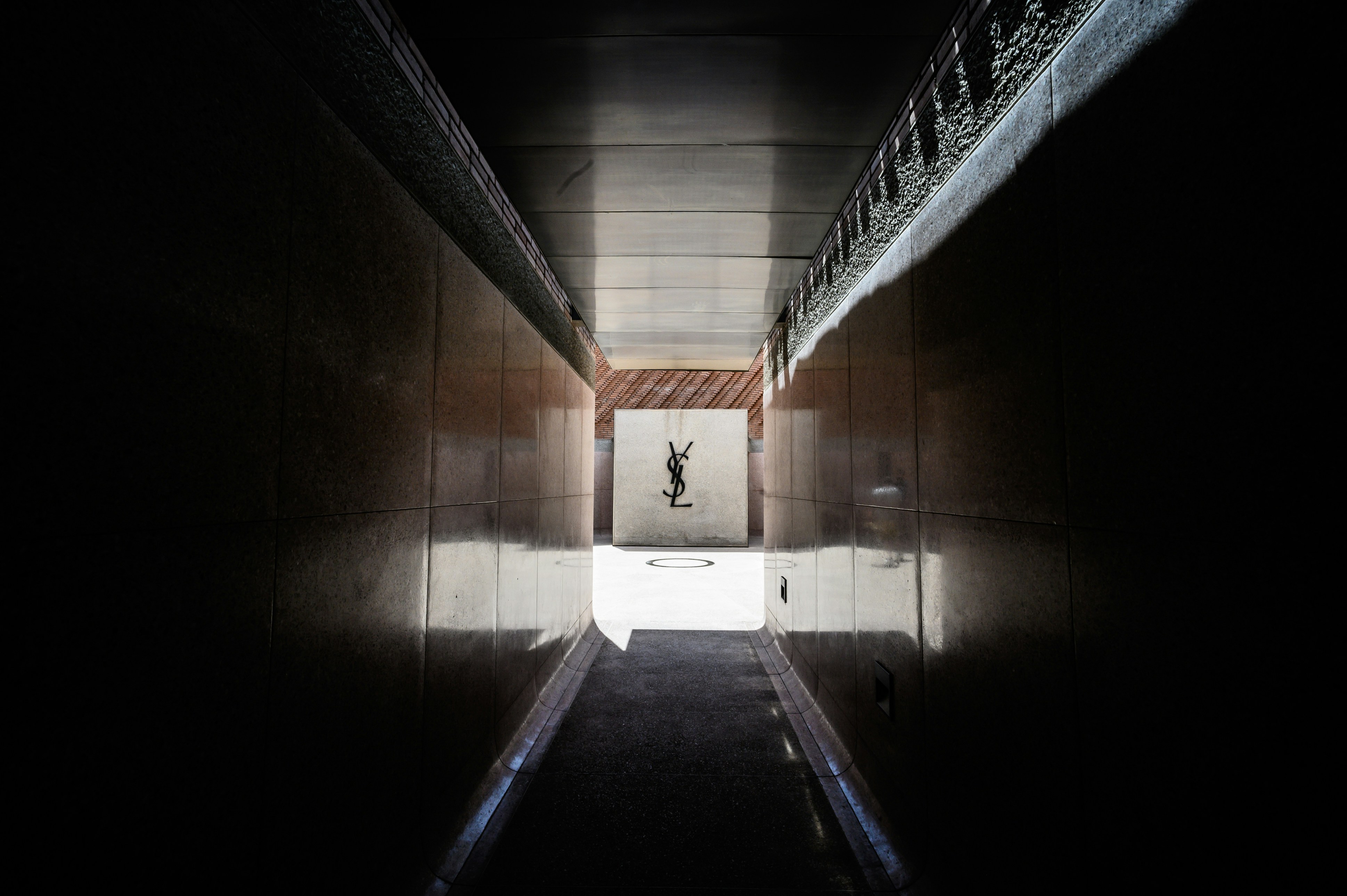 Dark passageway leading to a sunlit courtyard with a YSL logo on a stone wall.