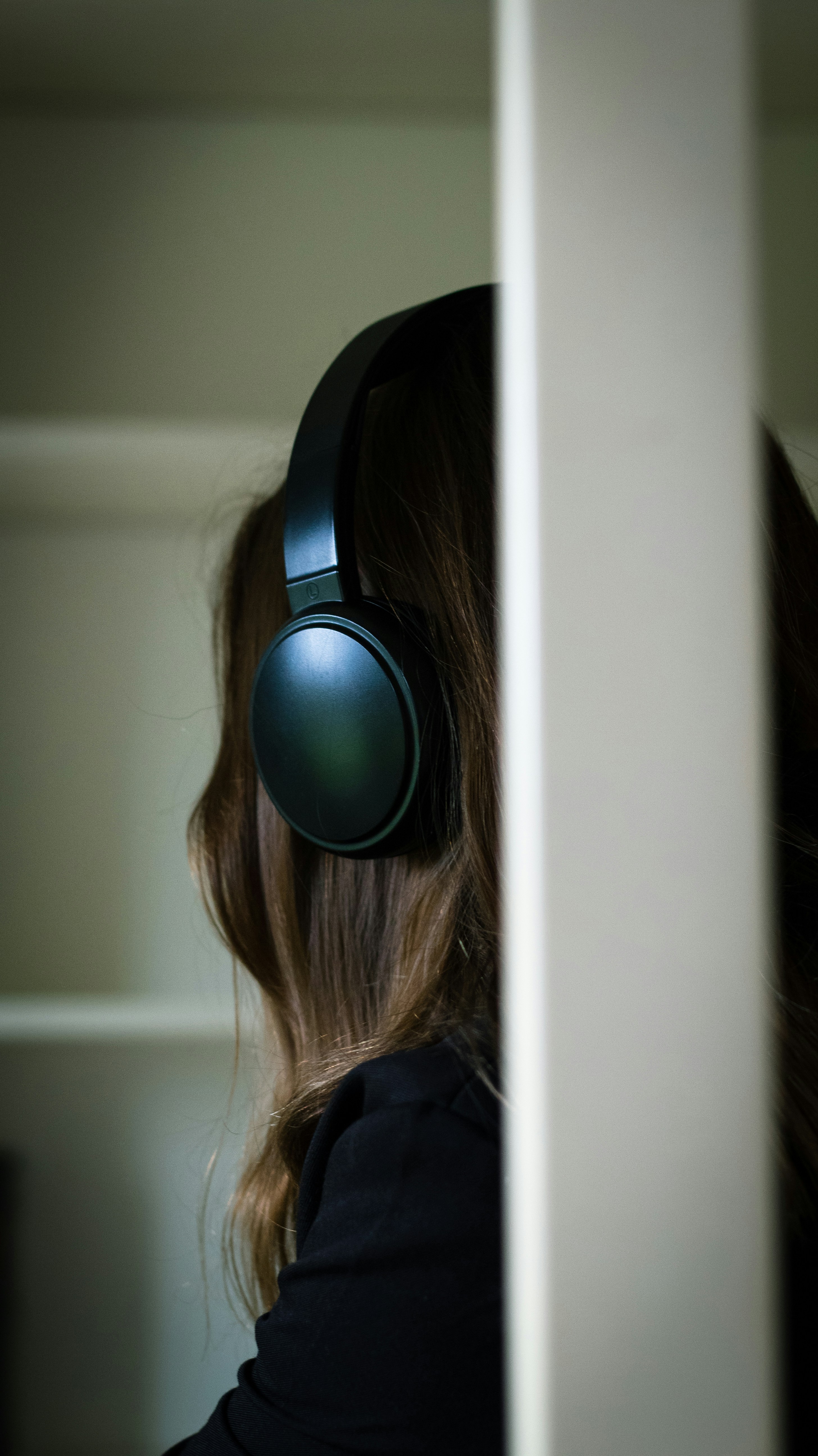 A woman with long hair wearing sleek black headphones, partially obscured by a white shelf, creating an intimate atmosphere of solitude.