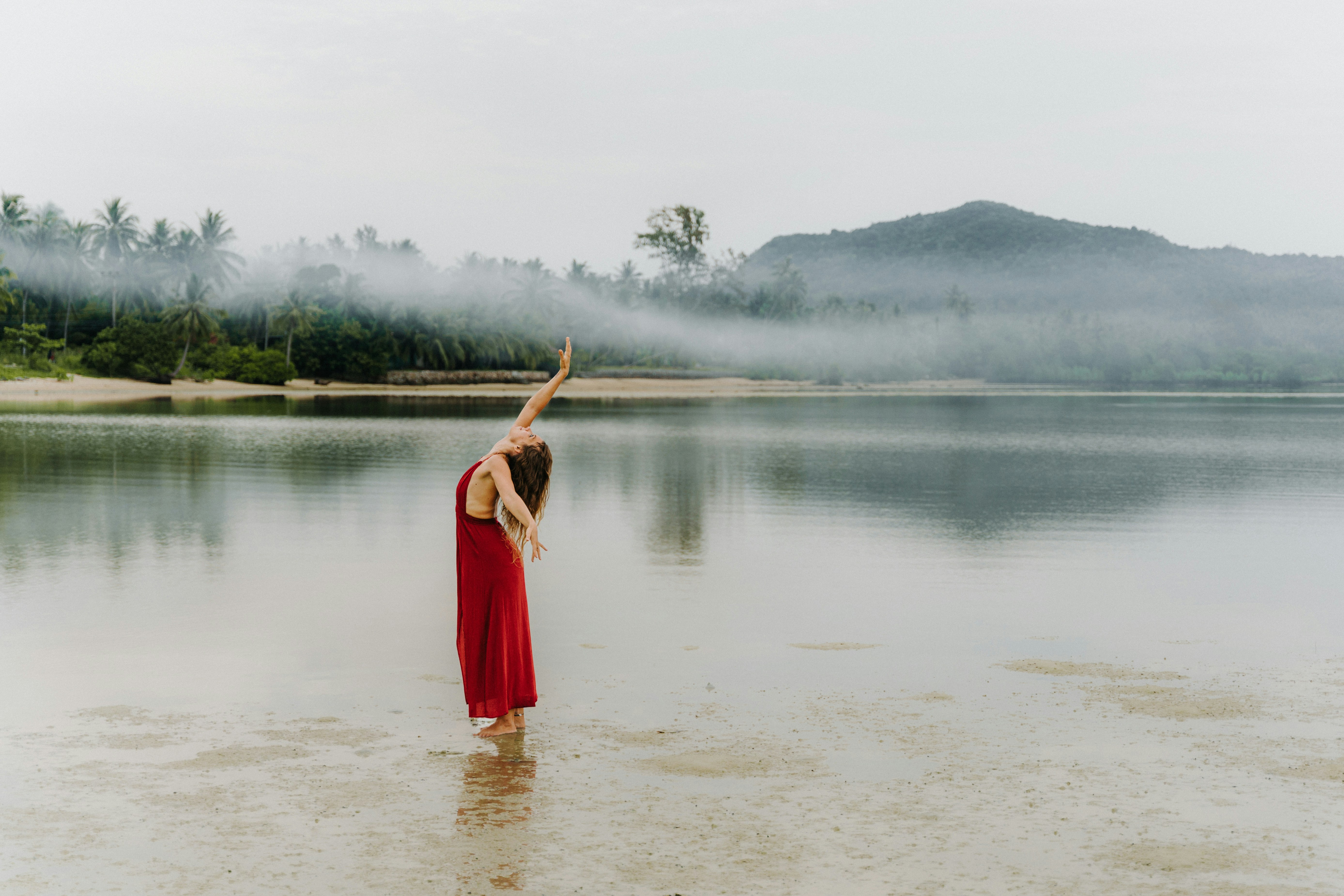 Person in a red dress stands on a tranquil lakeshore with misty mountains in the background.
