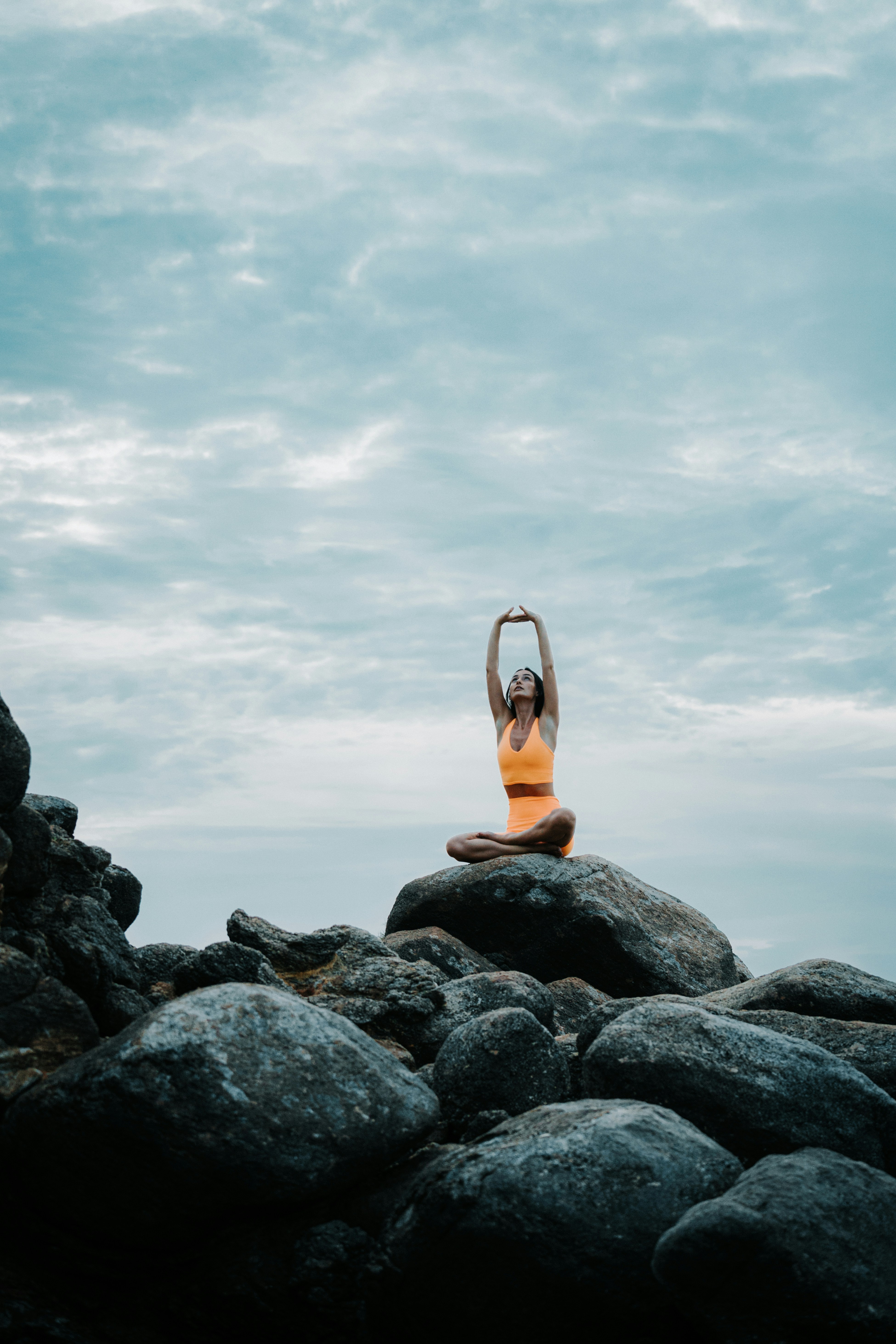 A woman in an orange swimsuit practices yoga atop a rocky outcrop, surrounded by a serene sky. The composition emphasizes tranquility and connection with nature.