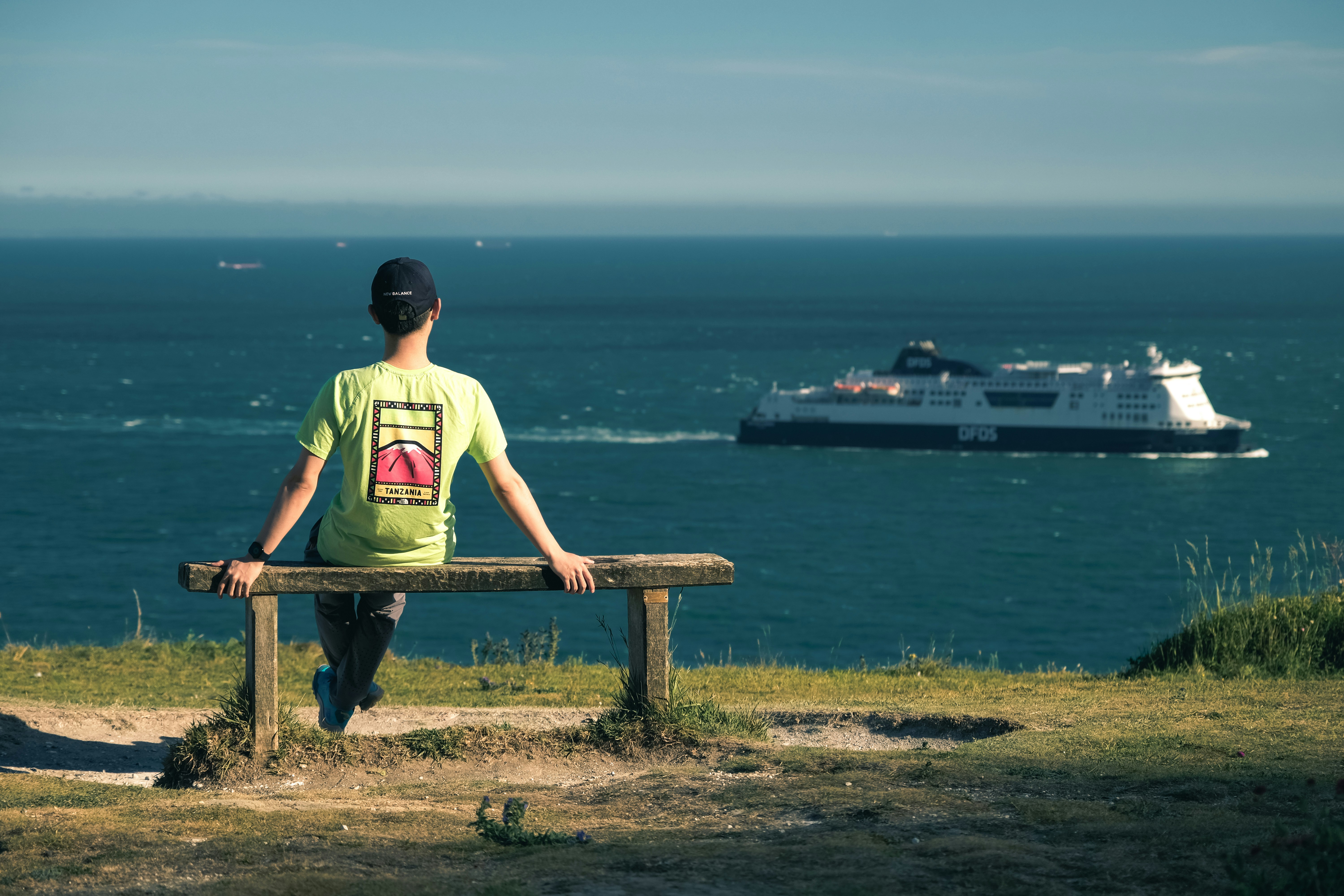A person in a bright green shirt sits on a bench overlooking a serene ocean, with a ferry passing in the distance.