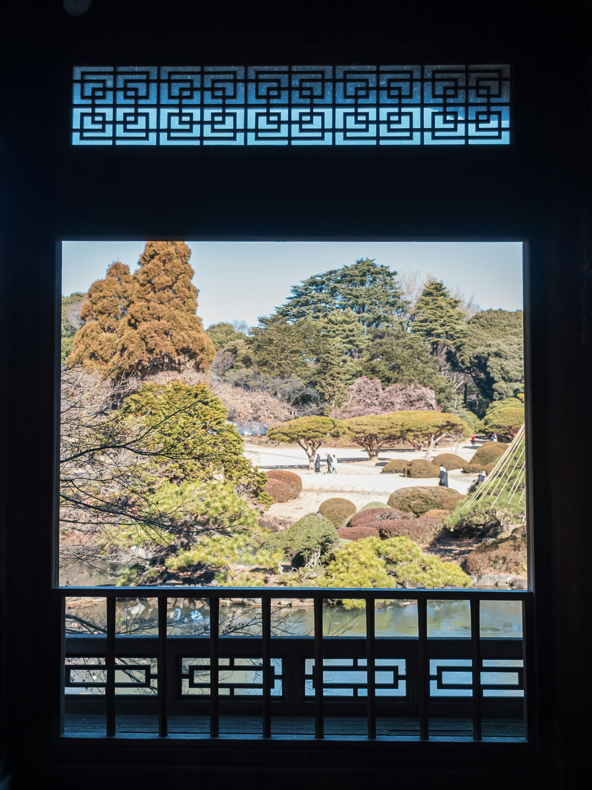 A photograph taken through a lattice-patterned window reveals a meticulously maintained Zen garden with trimmed trees, shrubs, and a tranquil pond. Distant visitors walk along a gravel path, adding scale.