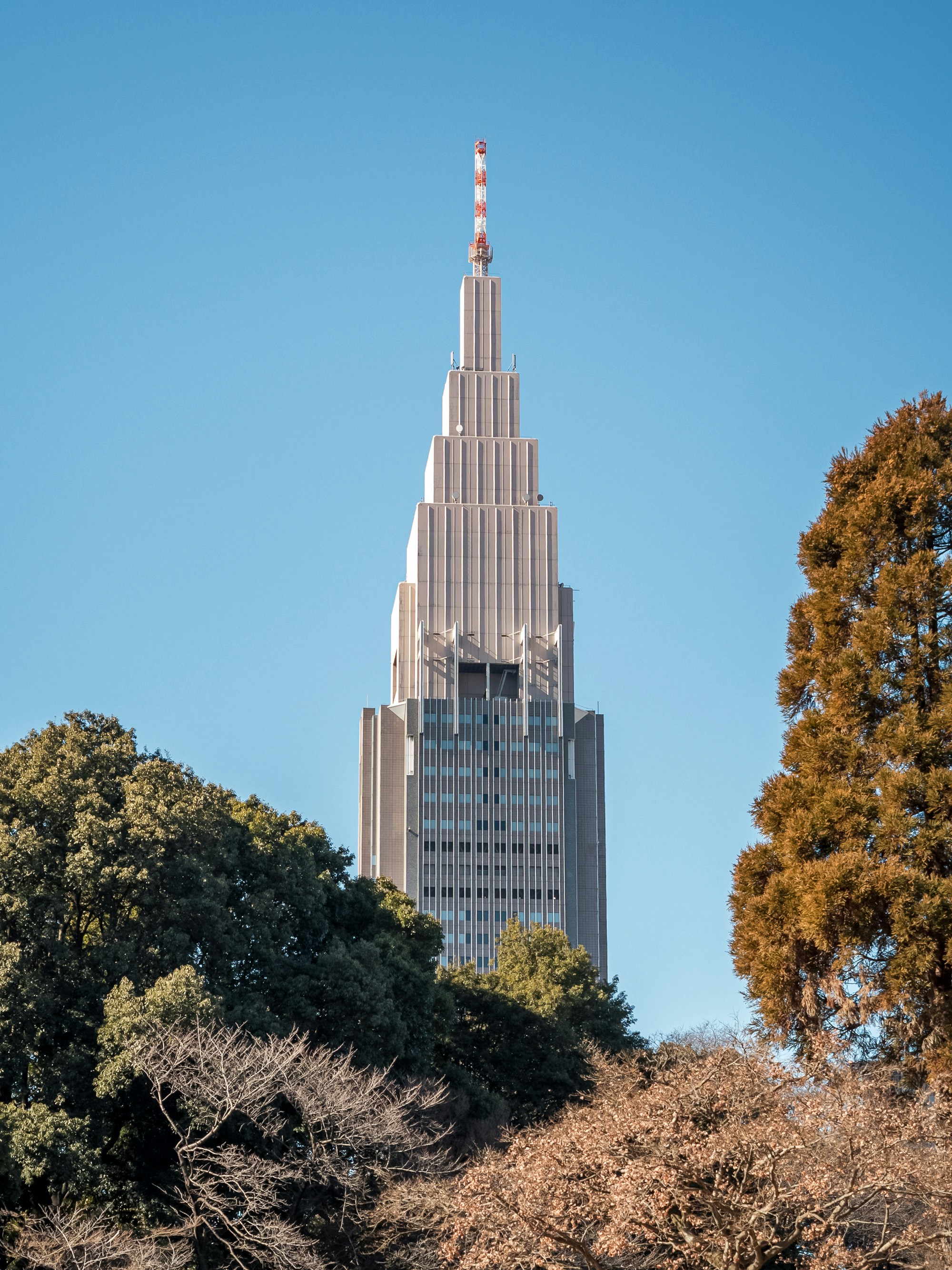 Tiered skyscraper rises above a tree line beneath a clear blue sky, its antenna crown projecting from the summit.