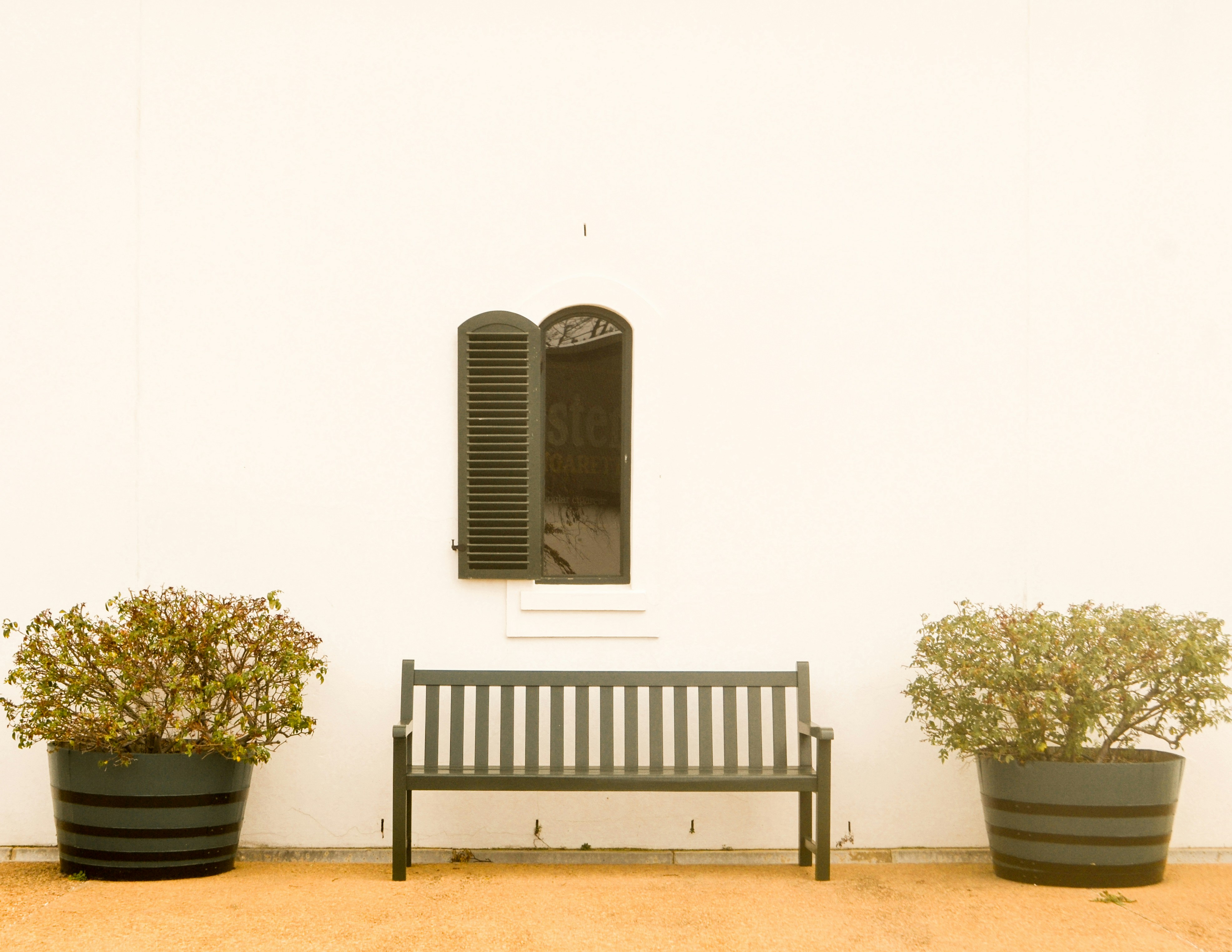 Black bench centered between two potted plants against a white wall with a single shuttered window above.