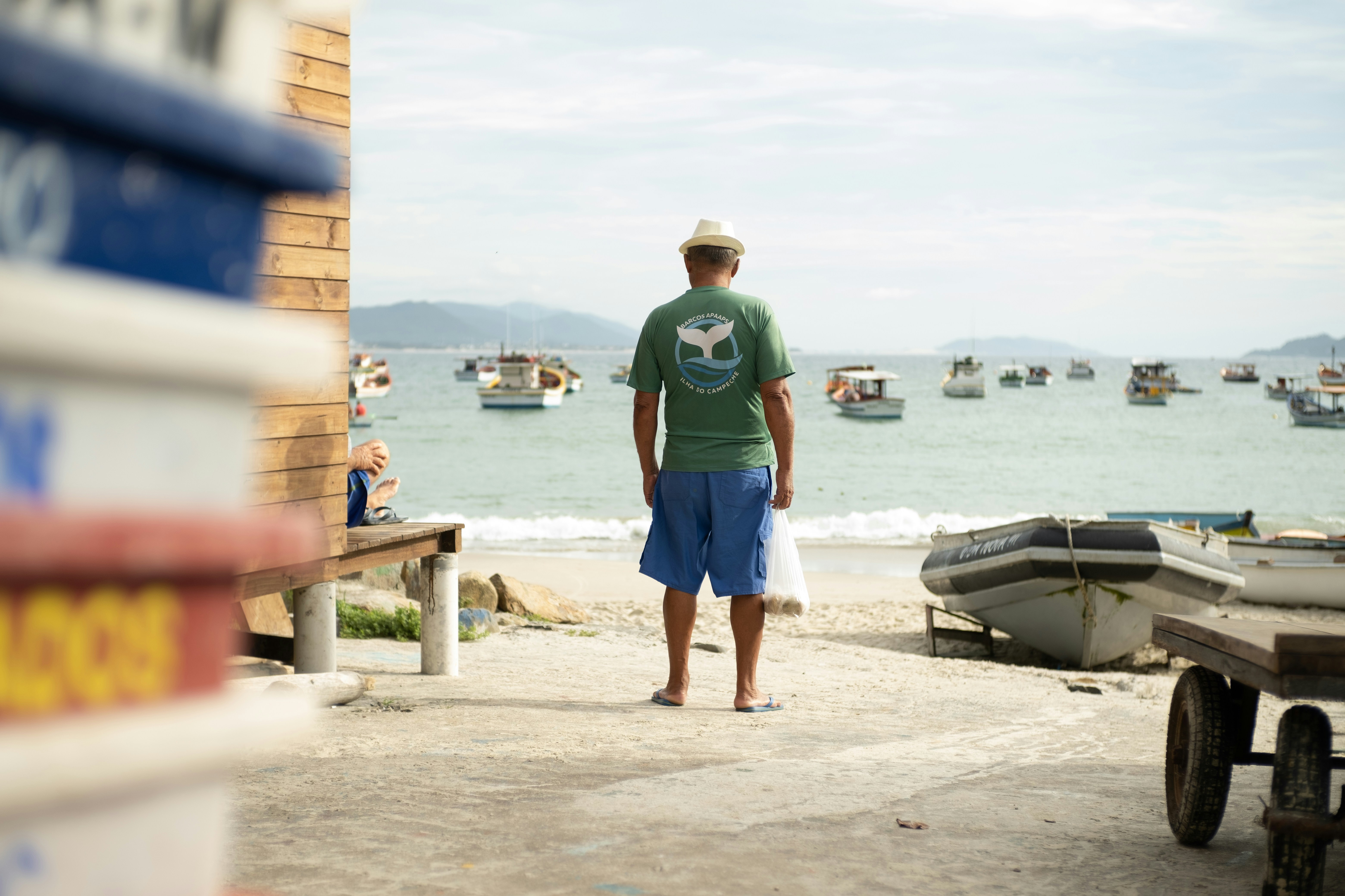 Man in hat and green shirt stands on a beach facing a fleet of boats on the ocean.