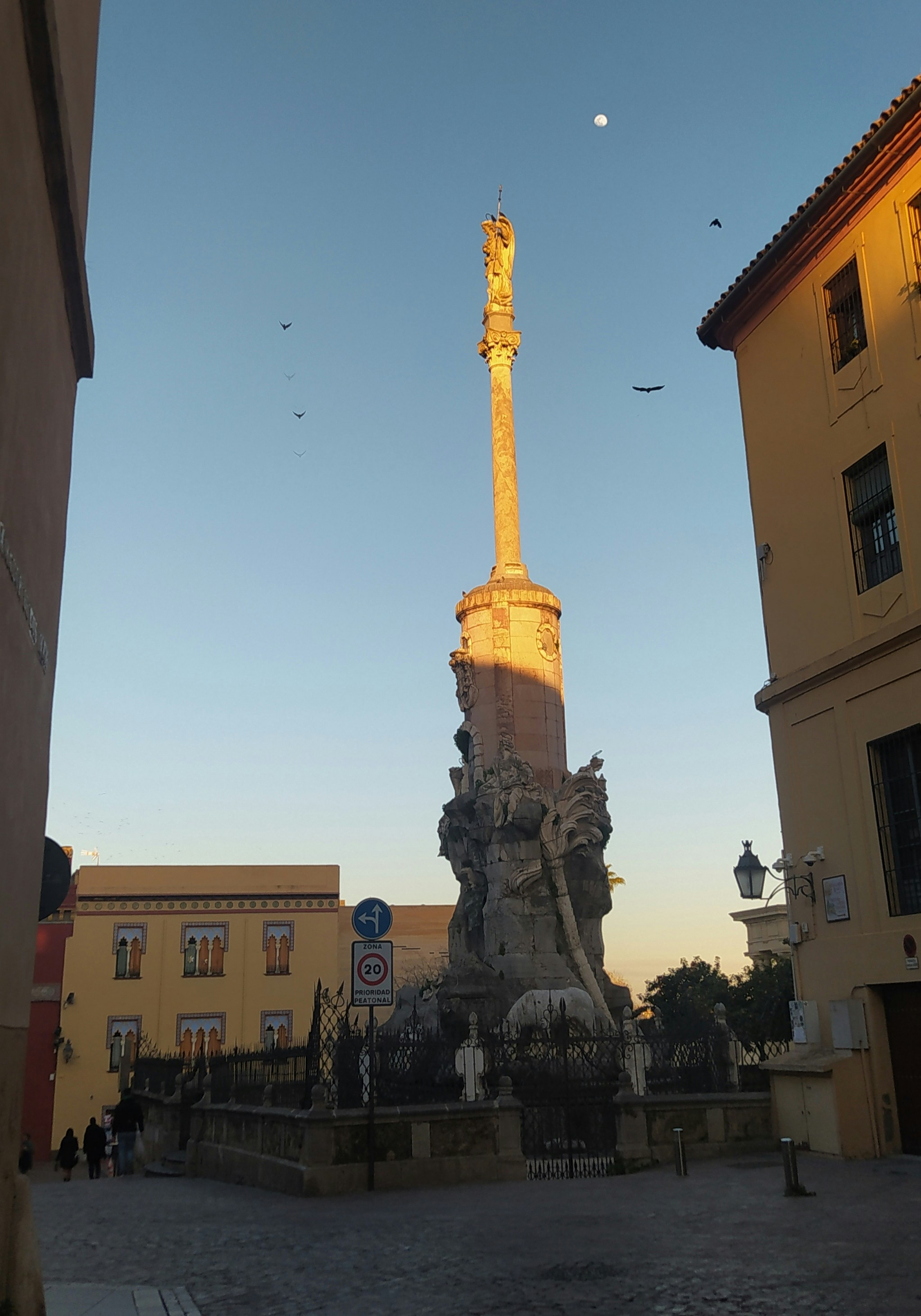 Illuminated monument surrounded by historic architecture under a clear evening sky, with a hint of the moon visible.