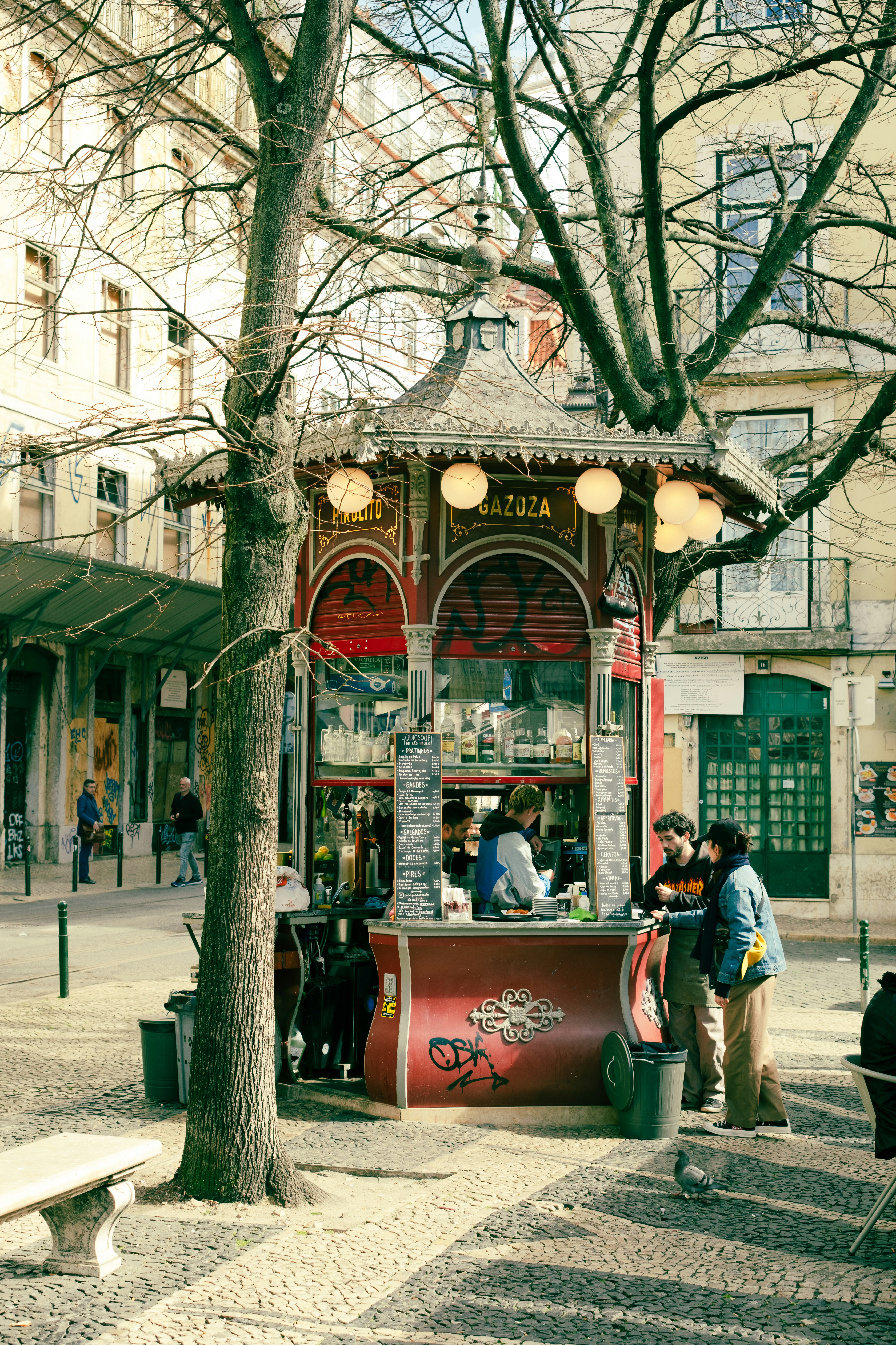 Charming kiosk in a vibrant square, surrounded by trees and urban art, where people gather for drinks and conversation.