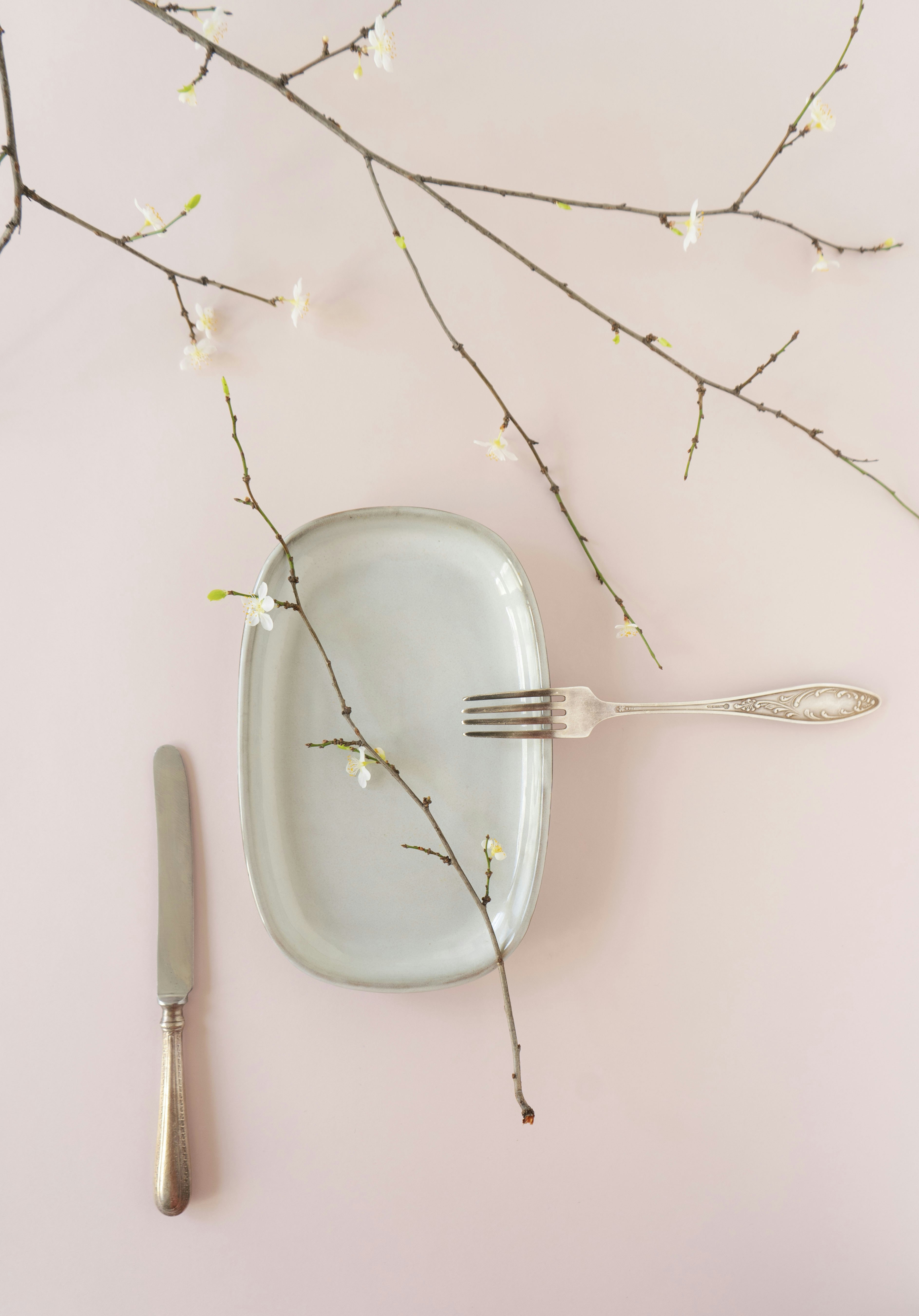 Delicate branches with blossoms artfully arranged over a pale plate and silverware on a soft pink background.