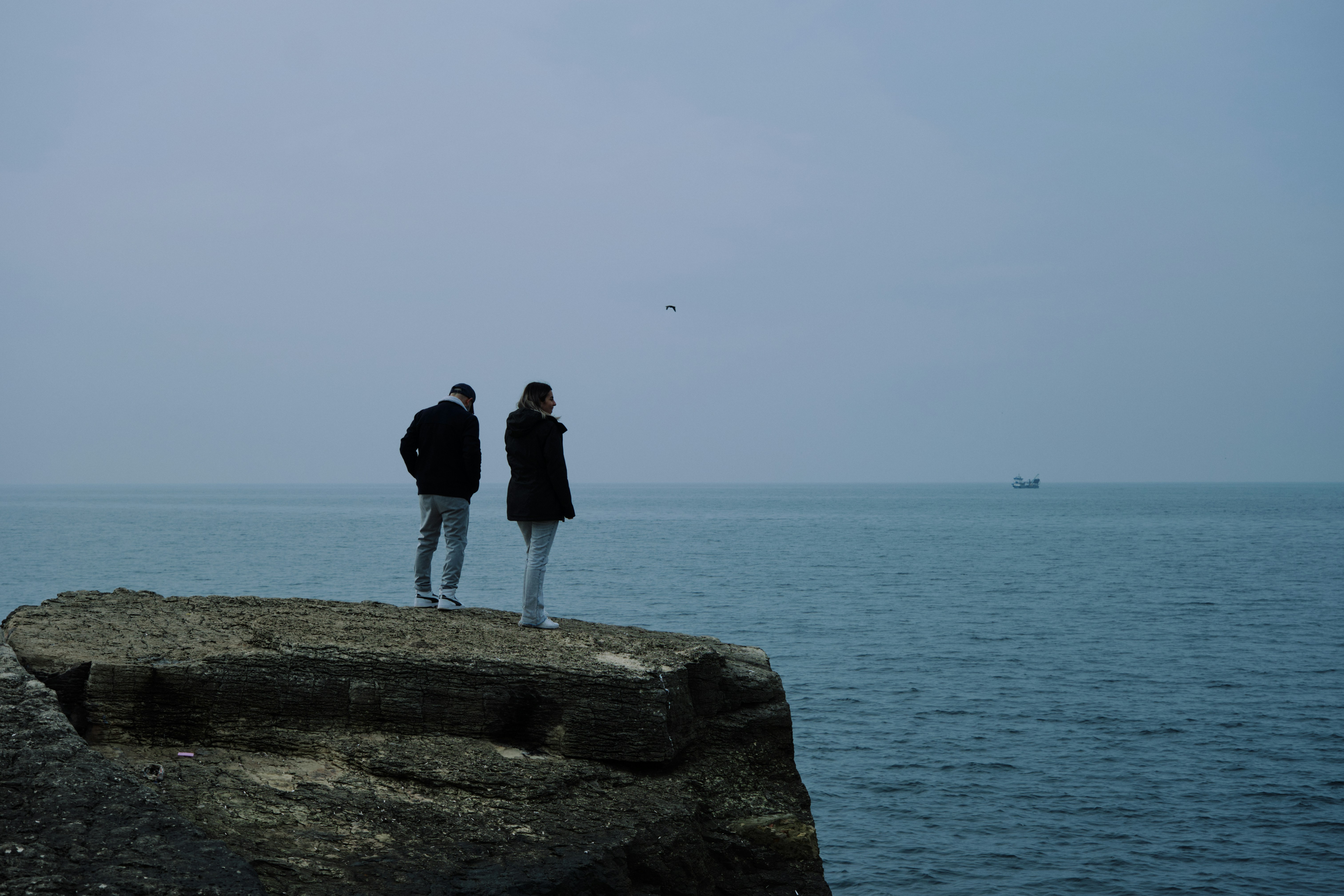 Two Silhouettes on a Cliff Edge Beneath a Pale Coastal Sky