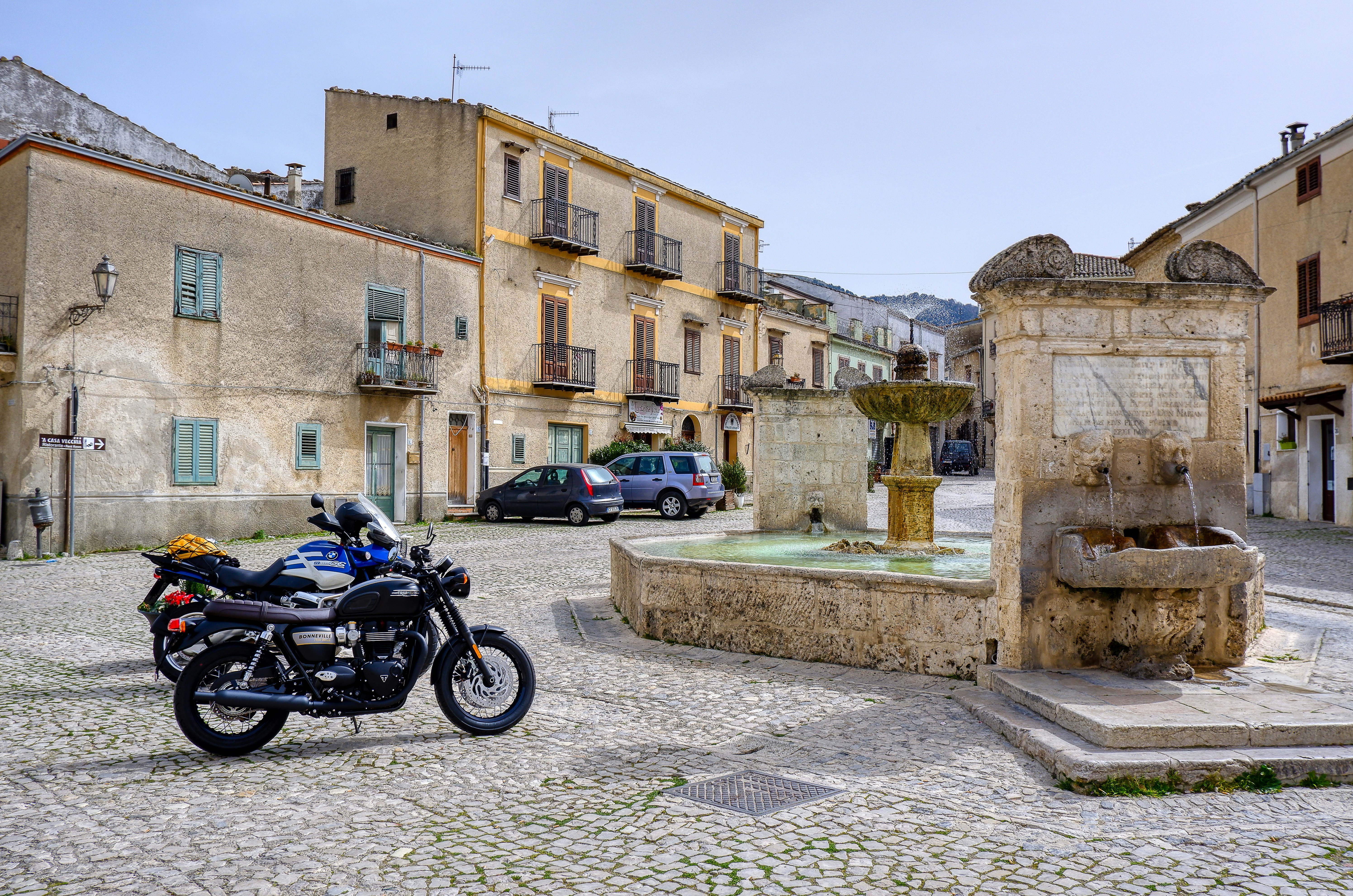 A parked motorcycle beside a historic fountain in a quaint village square, surrounded by rustic buildings and cobblestone streets.