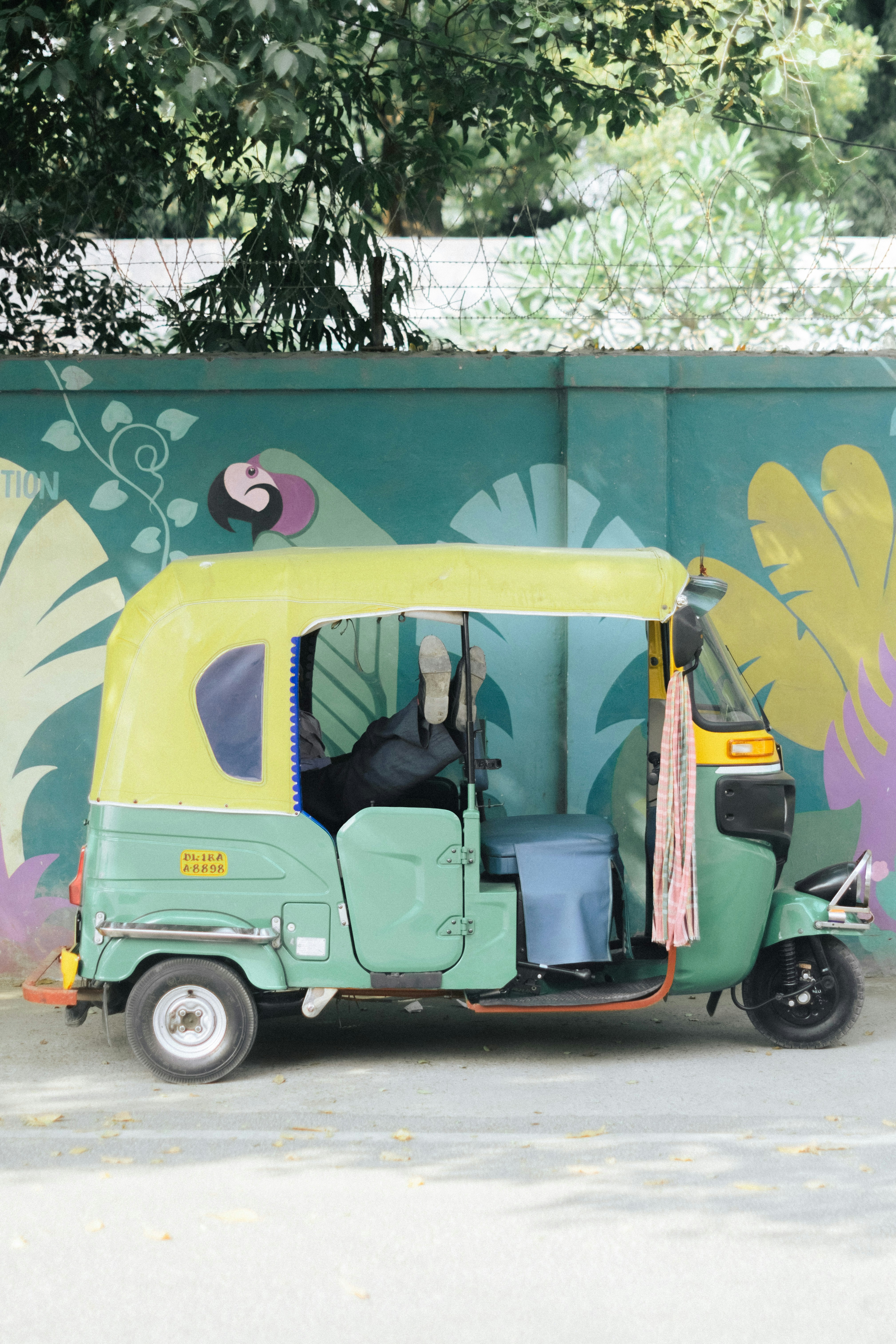 A green and yellow auto rickshaw parked against a vibrant mural, with a relaxed passenger reclining inside. The scene captures urban life and local culture.