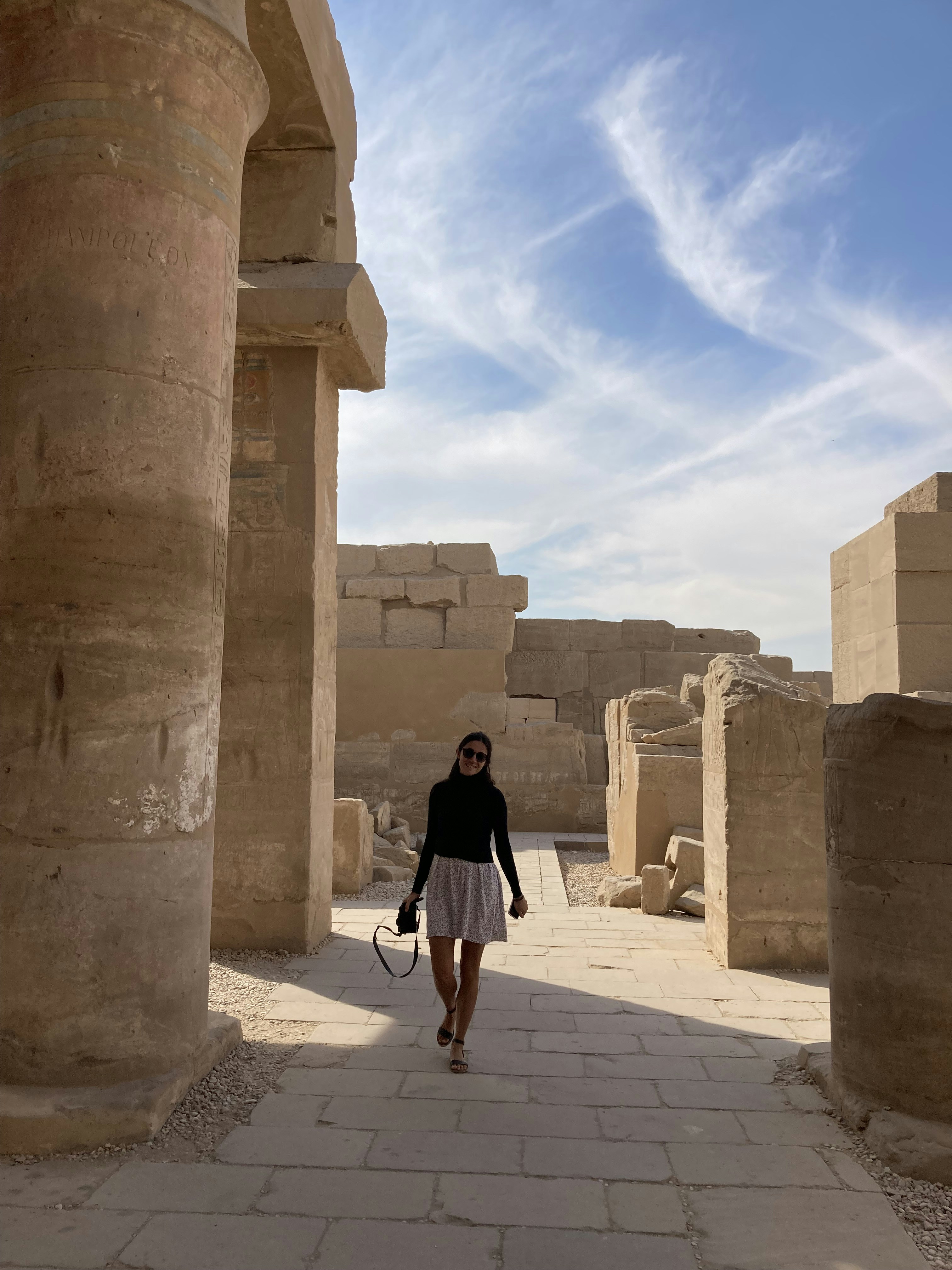 A woman walks through the ruins of an ancient structure, framed by towering columns and remnants of stone walls. The clear sky adds depth to the historic setting.