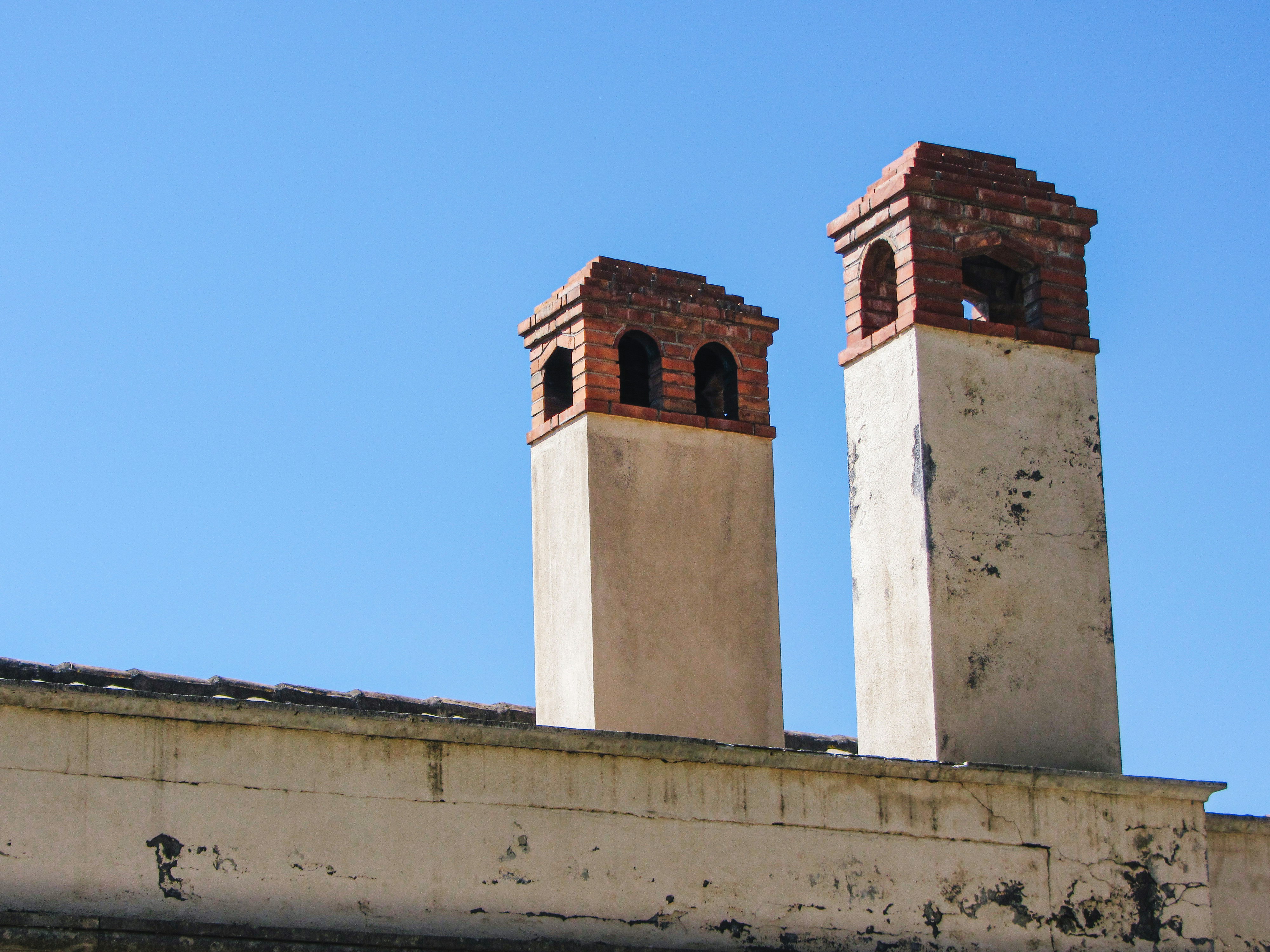 Two weathered brick chimneys rise against a clear blue sky, showcasing a blend of aging materials and historical craftsmanship.