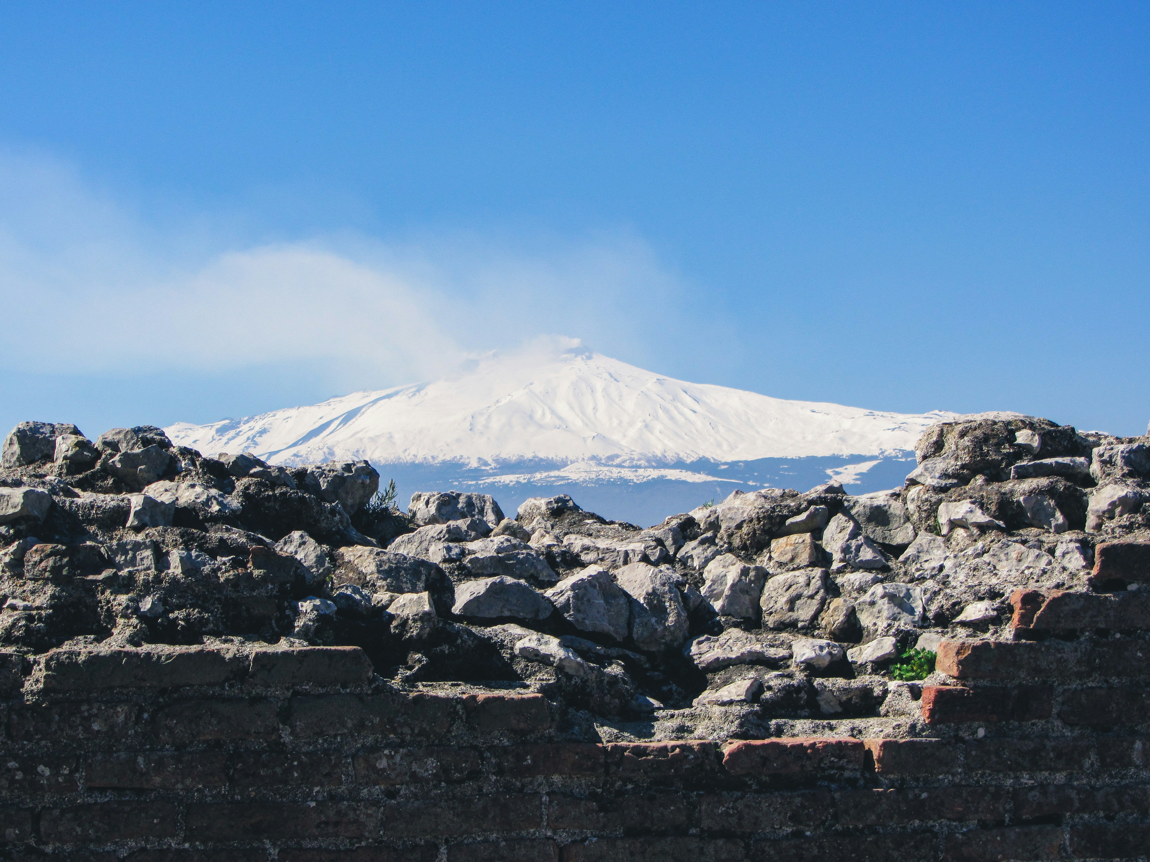 Snow-covered Mount Etna looms in the background, framed by ancient stone ruins, showcasing a blend of natural beauty and historical significance.