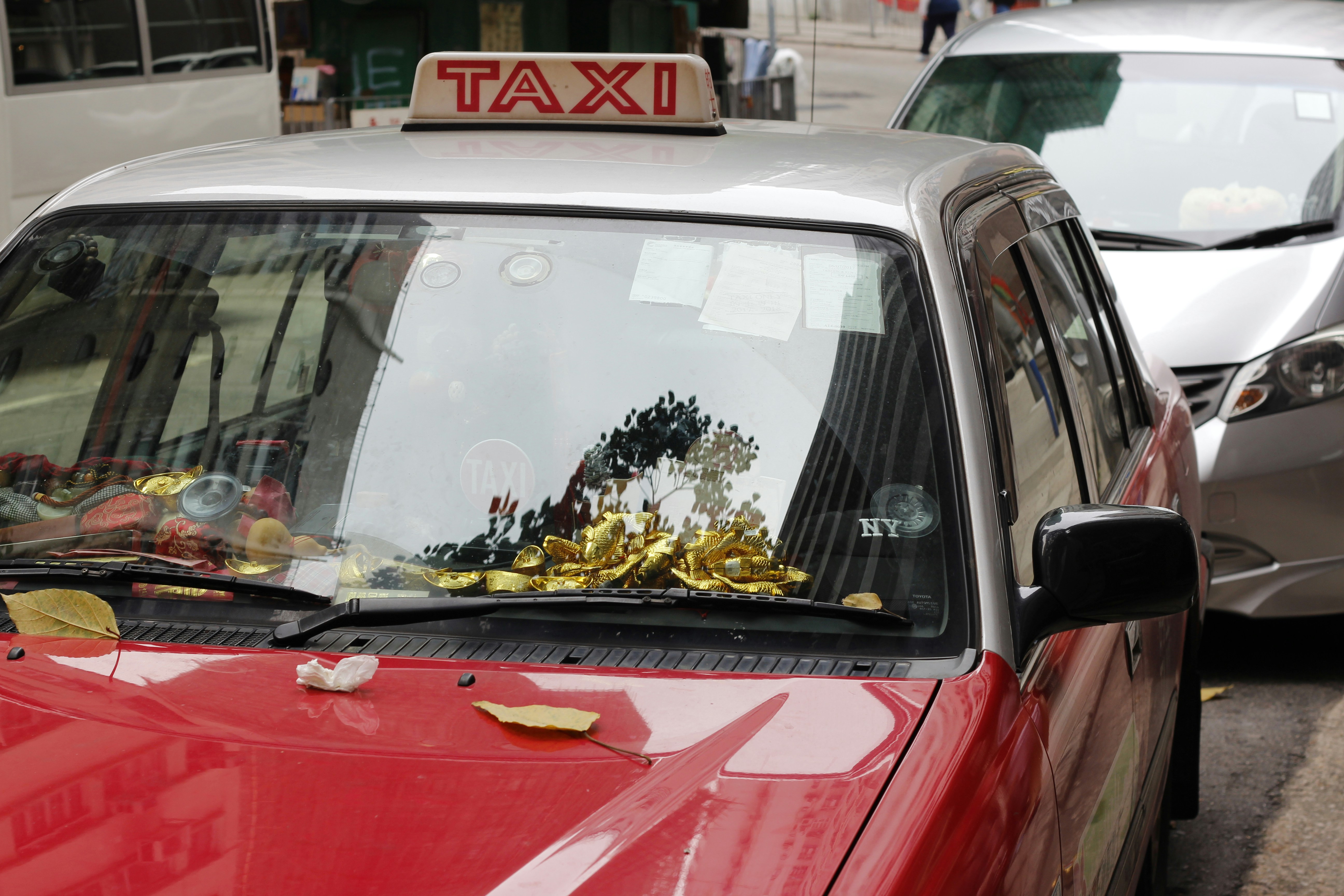 Red taxi parked on a city street, adorned with fallen leaves on the windshield and hood, reflecting a quiet urban scene.