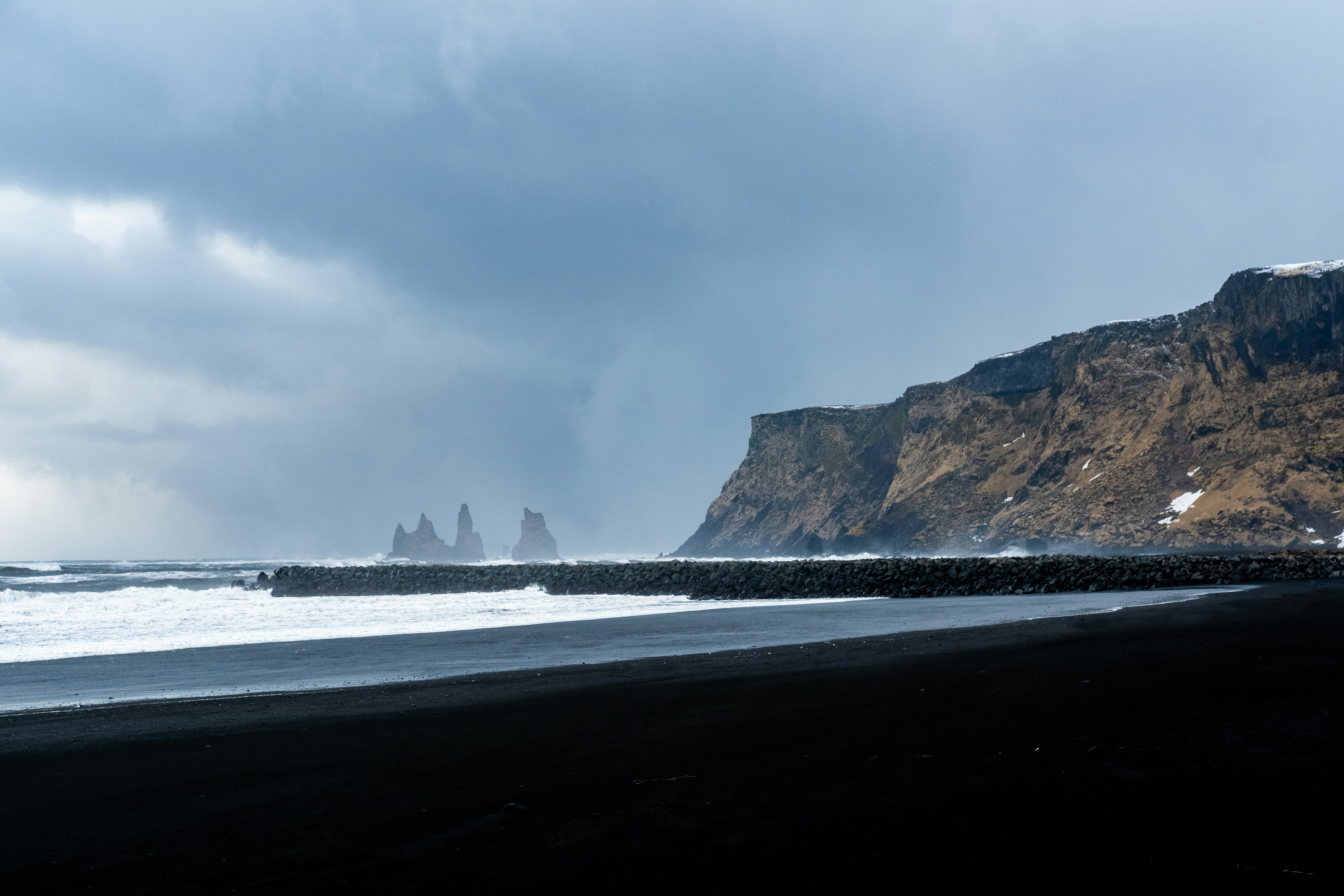 Dramatic coastal cliffs under a cloudy sky with dark sand beach and distant sea stacks.